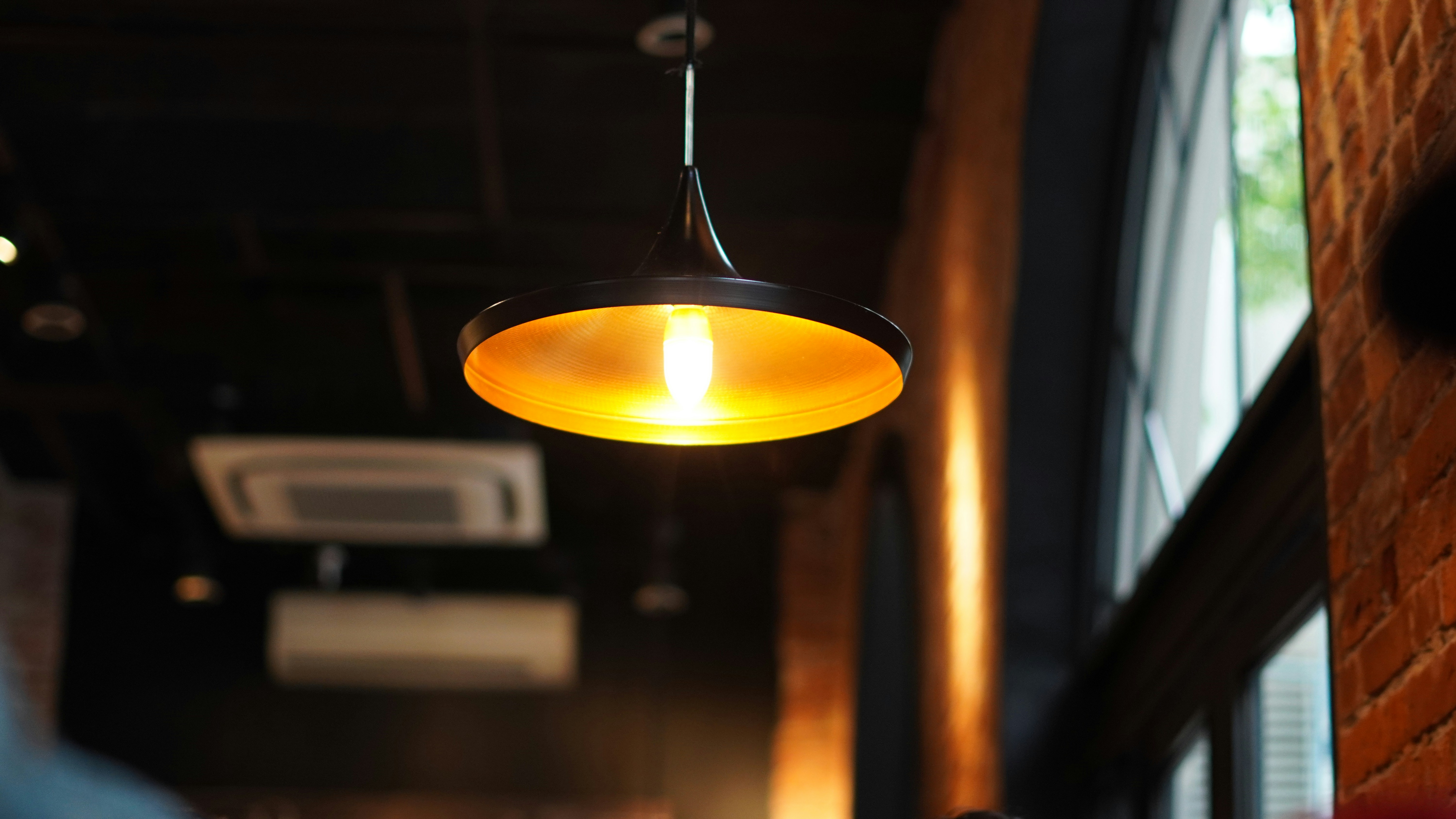 A stylish pendant lamp casts a warm glow in a cozy cafe setting, highlighting the interplay of light and shadow. The brick wall adds texture to the ambiance.