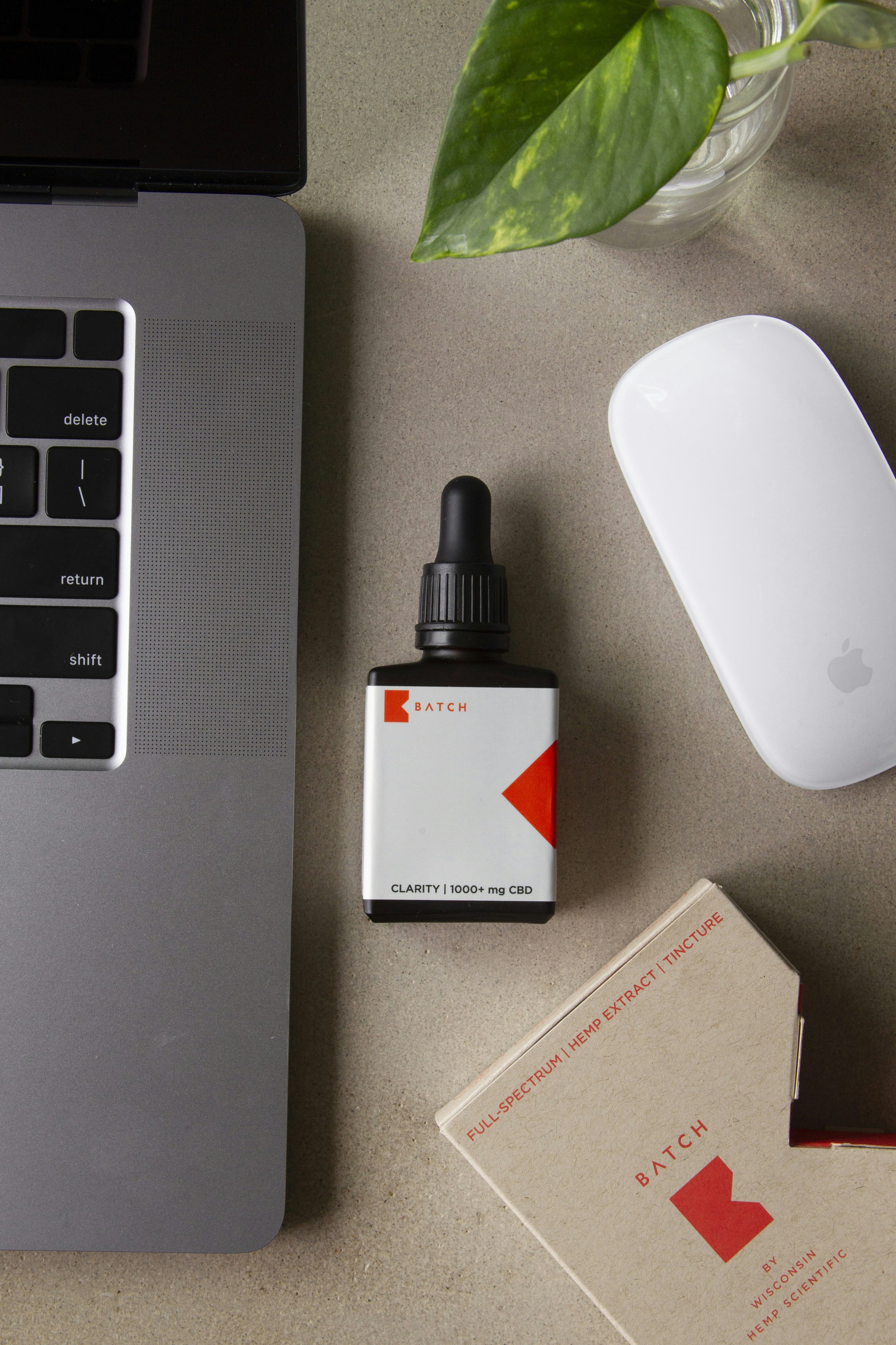 A desk scene featuring a laptop, a dropper bottle labeled CBD, a white wireless case, and a textured box, arranged on a neutral surface for product-focused photography.