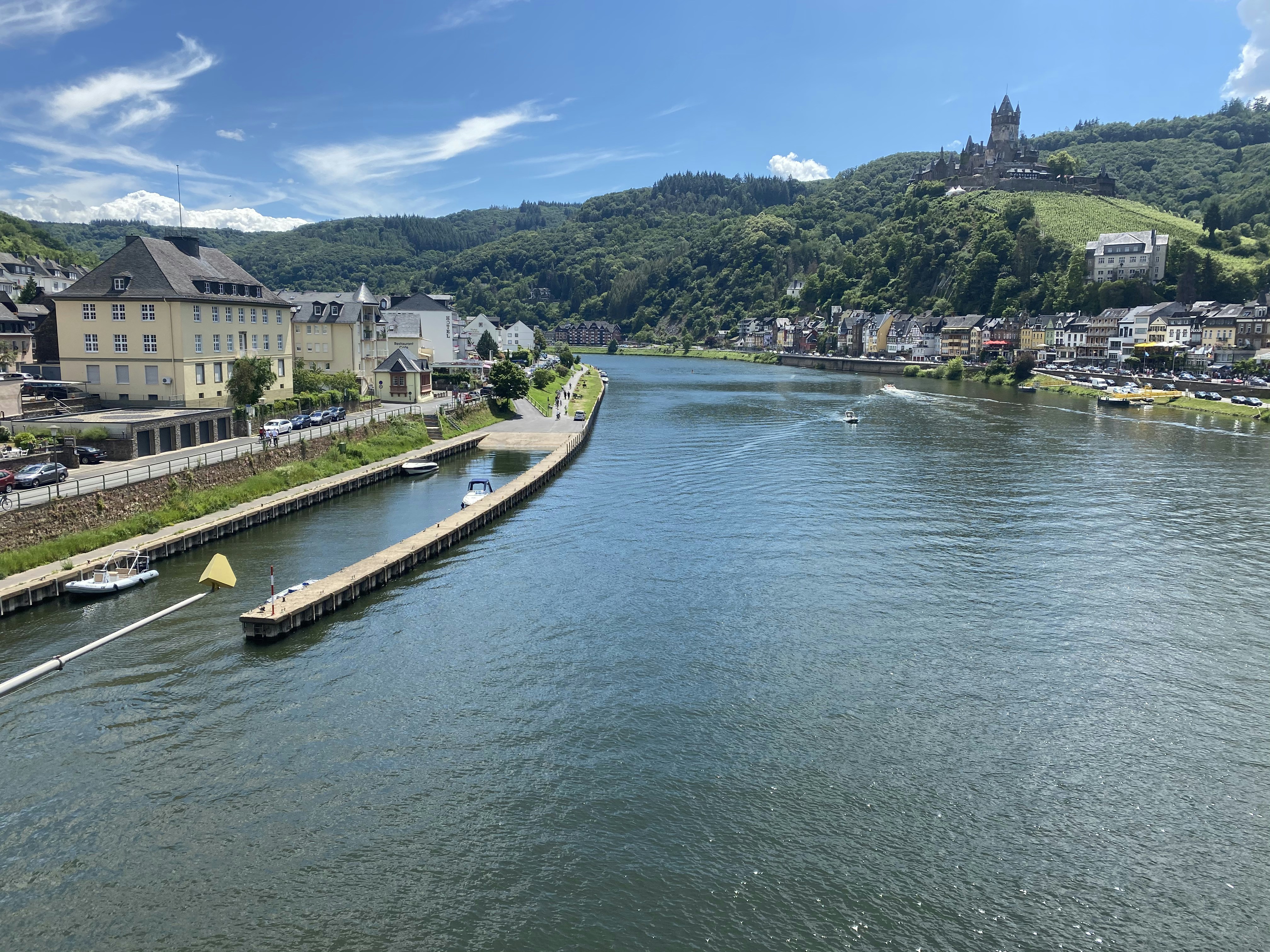 A tranquil river scene featuring boats along the shore, with lush hills and a castle in the background under a bright blue sky.