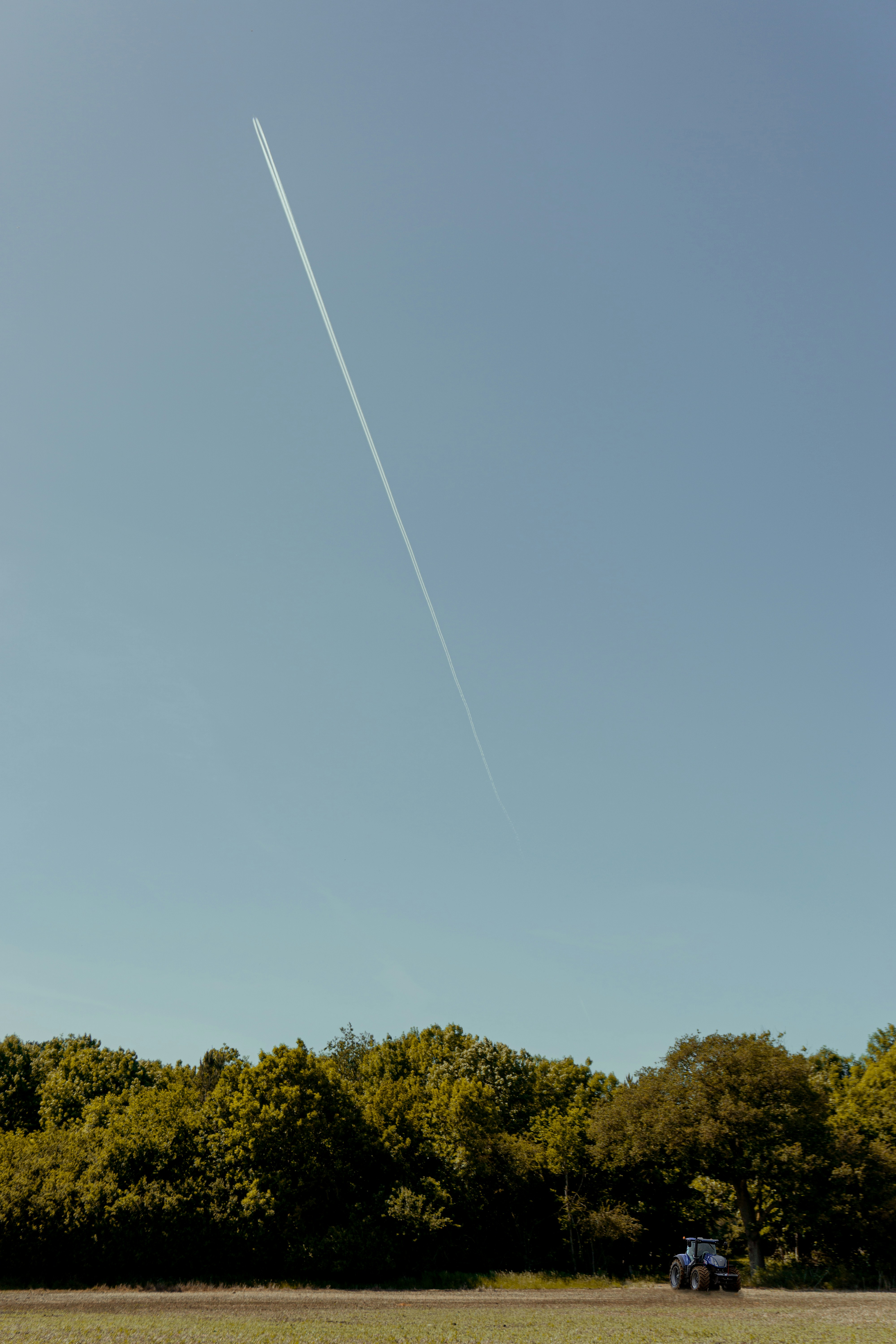 A solitary tractor in a field beneath a clear blue sky, marked by a distinct contrail stretching across the horizon.