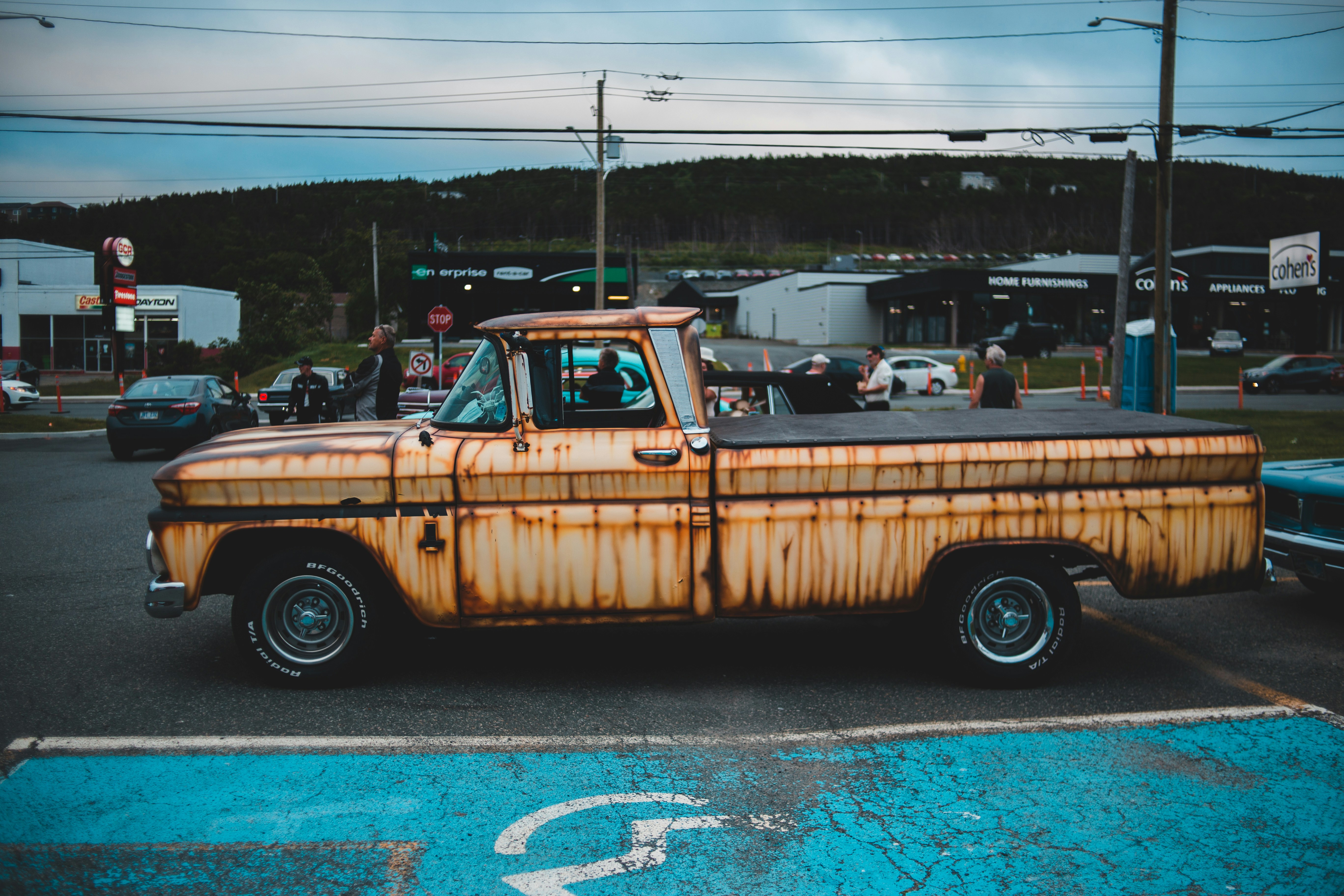 Weathered vintage pickup truck parked in a lot, showcasing its rustic patina and classic design. The backdrop features a bustling scene with other vehicles and buildings.
