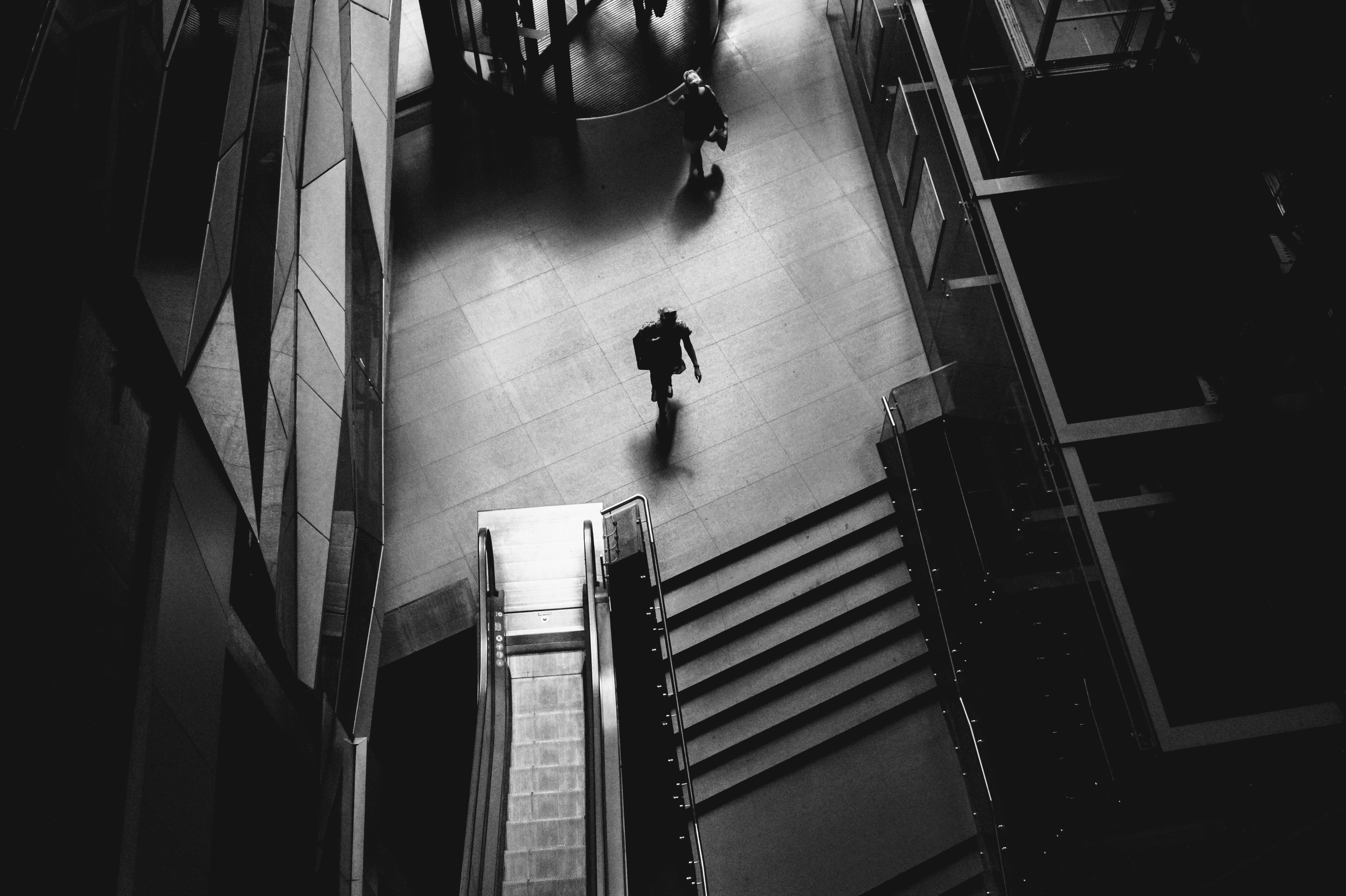 Black and white overhead shot of a person walking through a modern architectural space with stairs and escalators.
