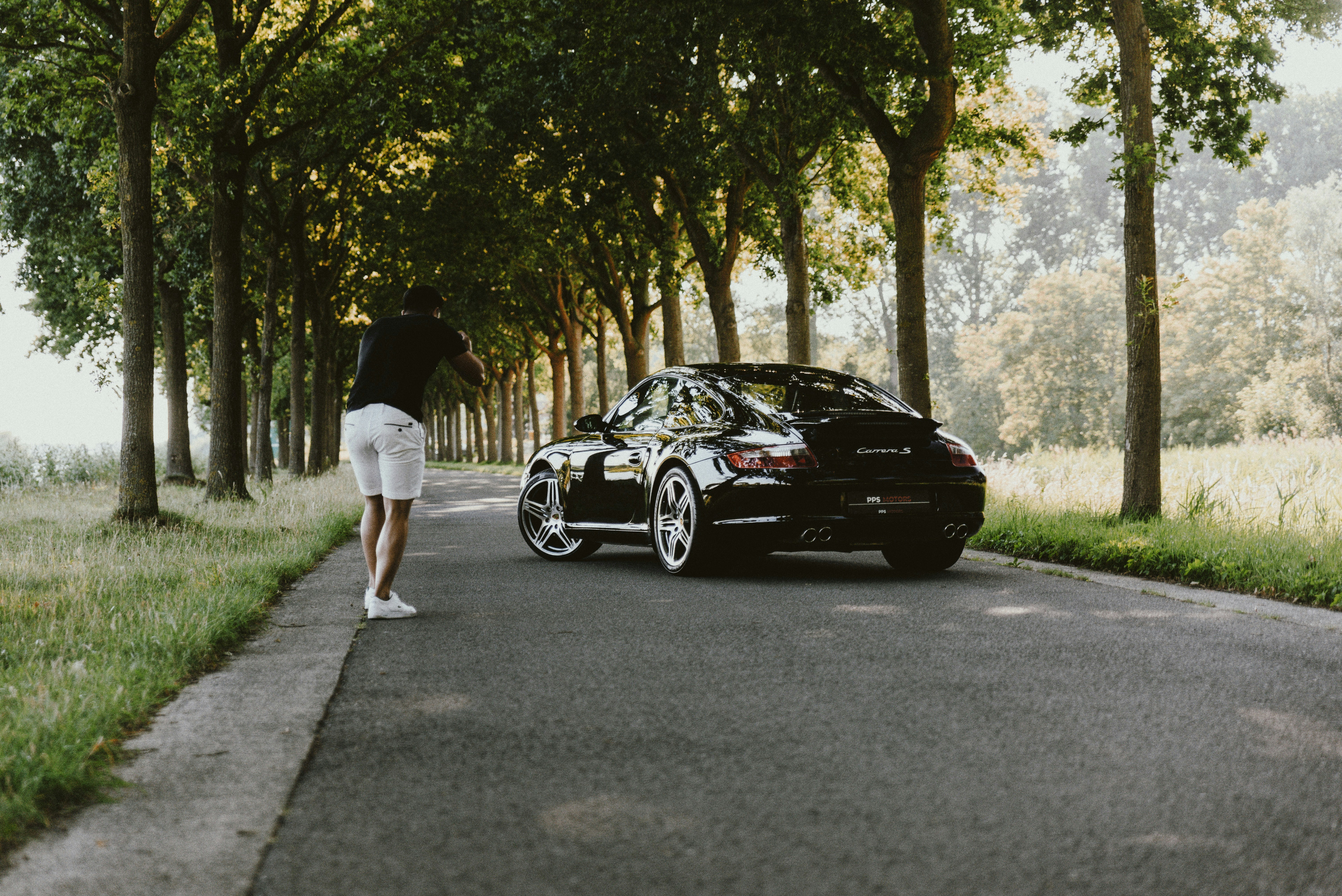 A sleek black sports car parked on a tree-lined avenue, with a photographer capturing its essence. The scene exudes a sense of freedom and adventure.