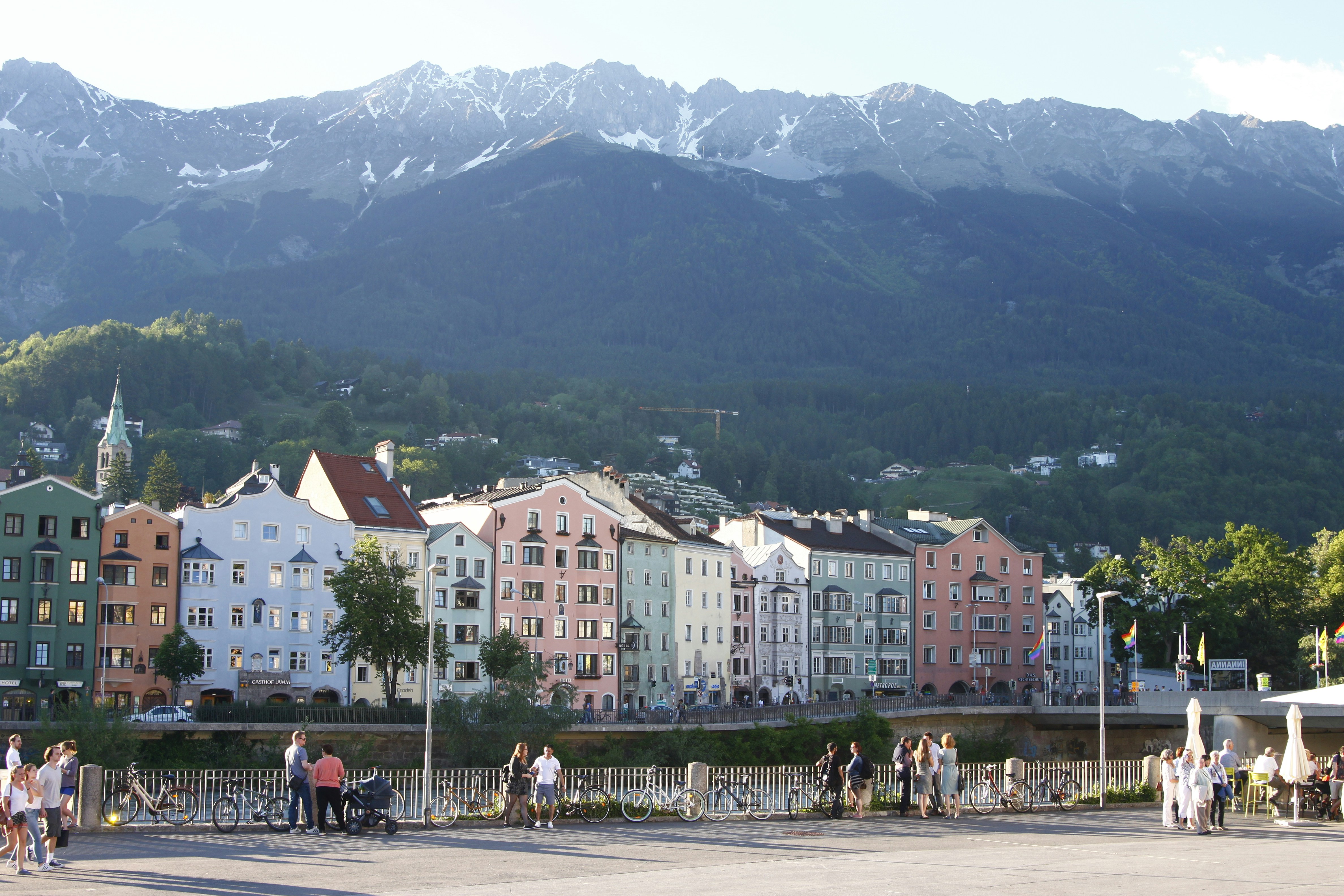 Charming pastel-colored buildings line the riverbank, with snow-capped mountains towering in the background. The scene captures a lively atmosphere with people enjoying the outdoors.