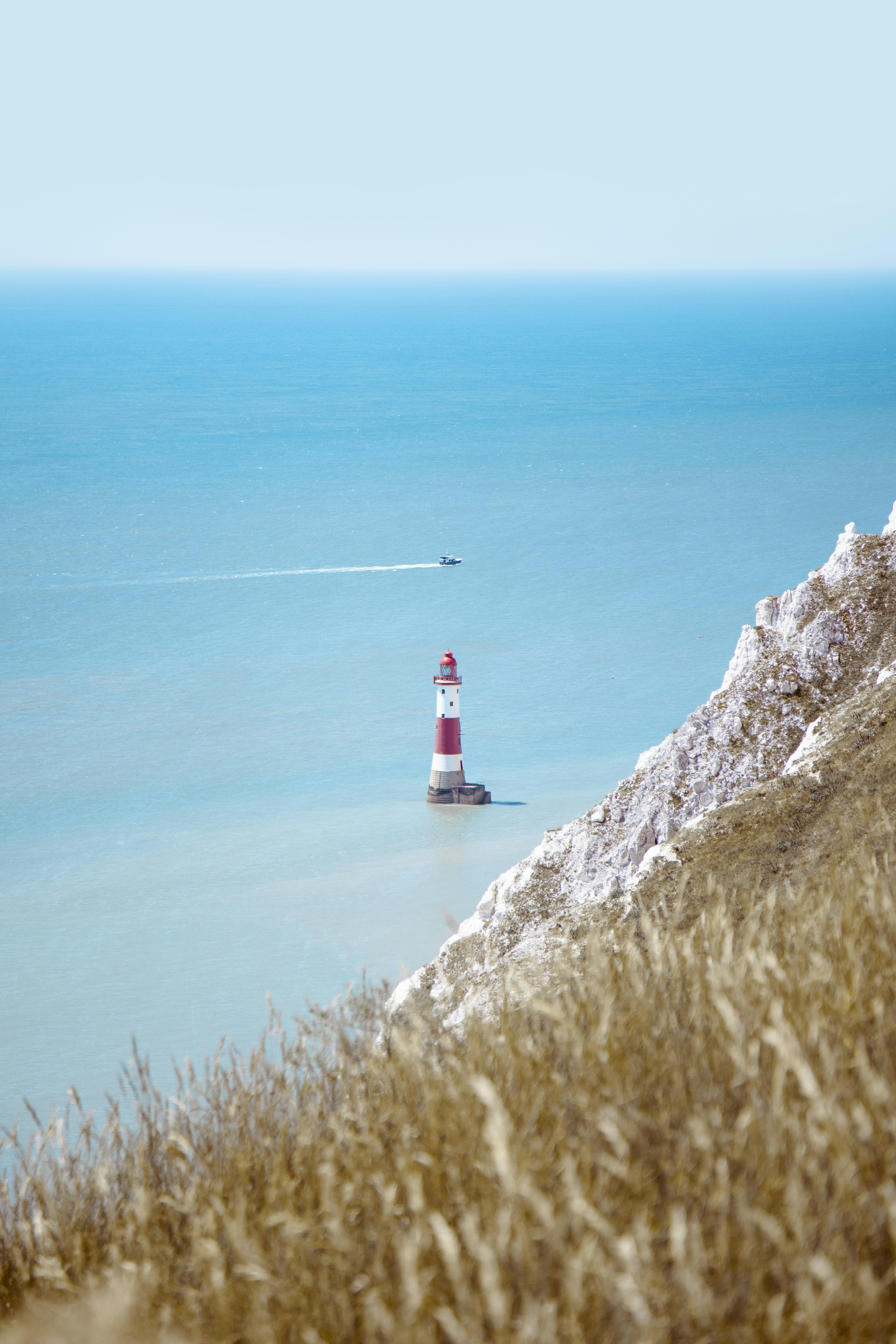 A solitary lighthouse stands tall in the serene waters, with a boat passing by, framed by a rugged cliff and gentle grasses in the foreground.