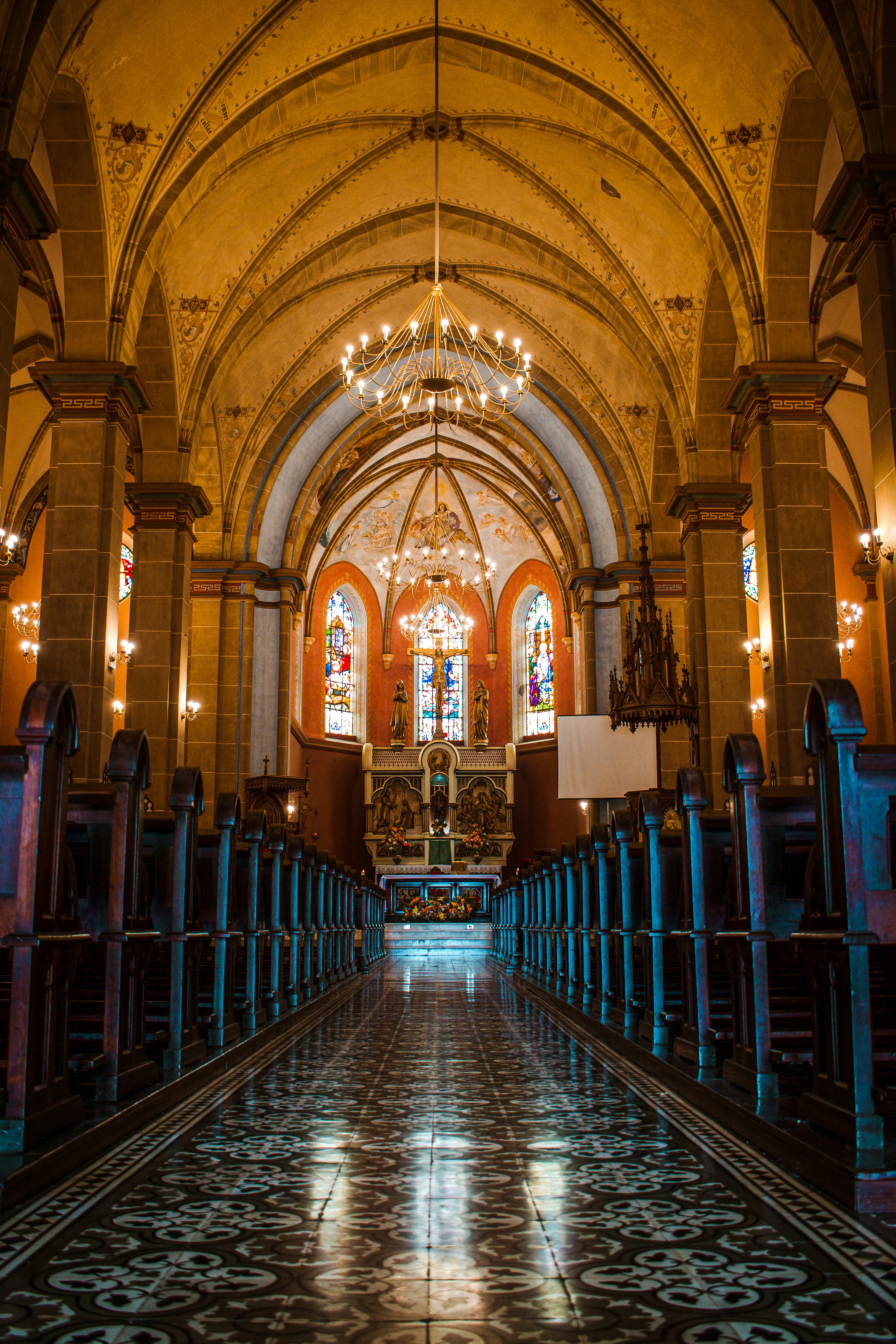 Ornate church interior with arched ceilings and intricate tile floor, illuminated by chandeliers.