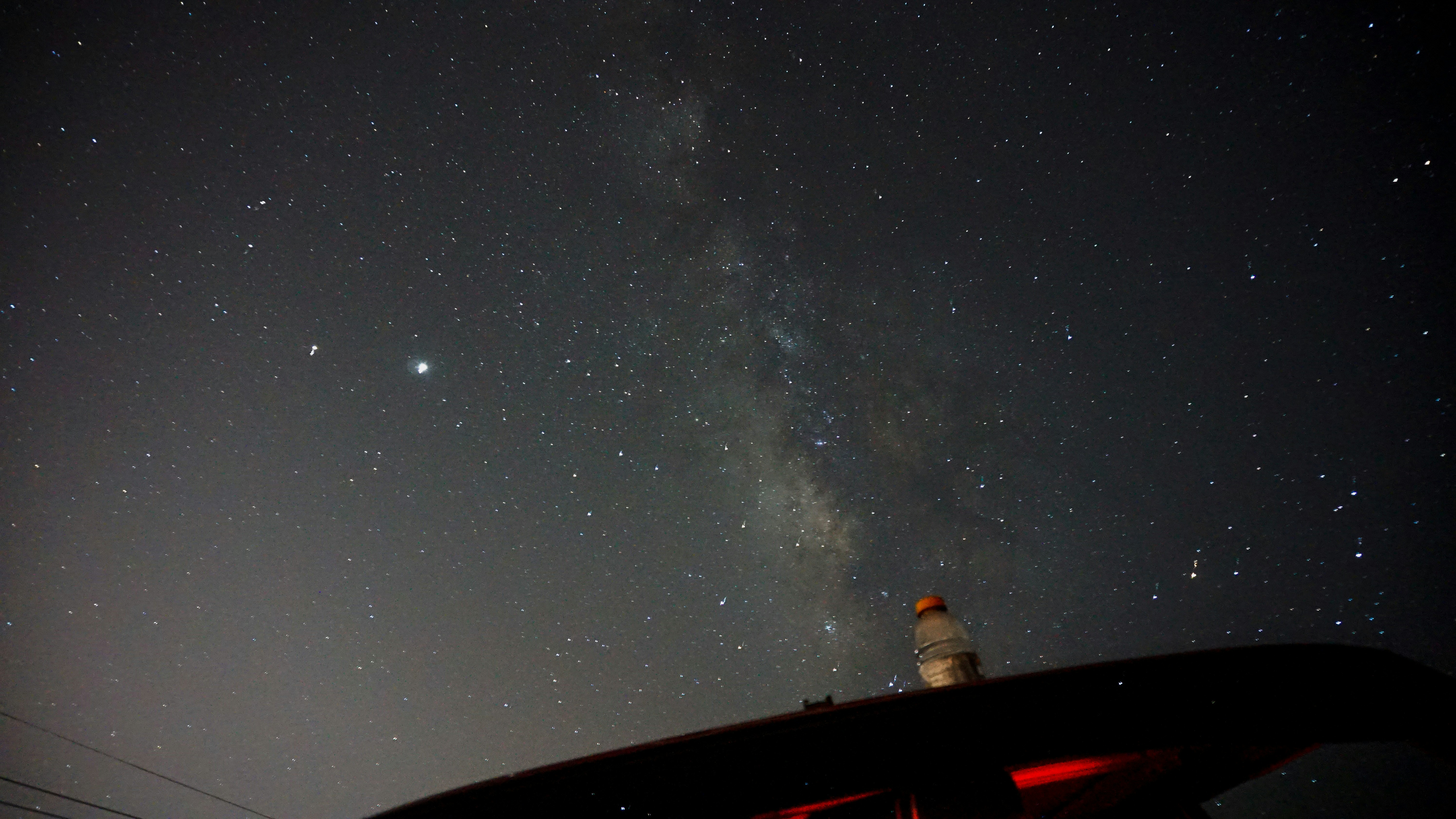 Milky Way galaxy illuminating the night sky with a silhouette of a bottle and vehicle roof in the foreground.