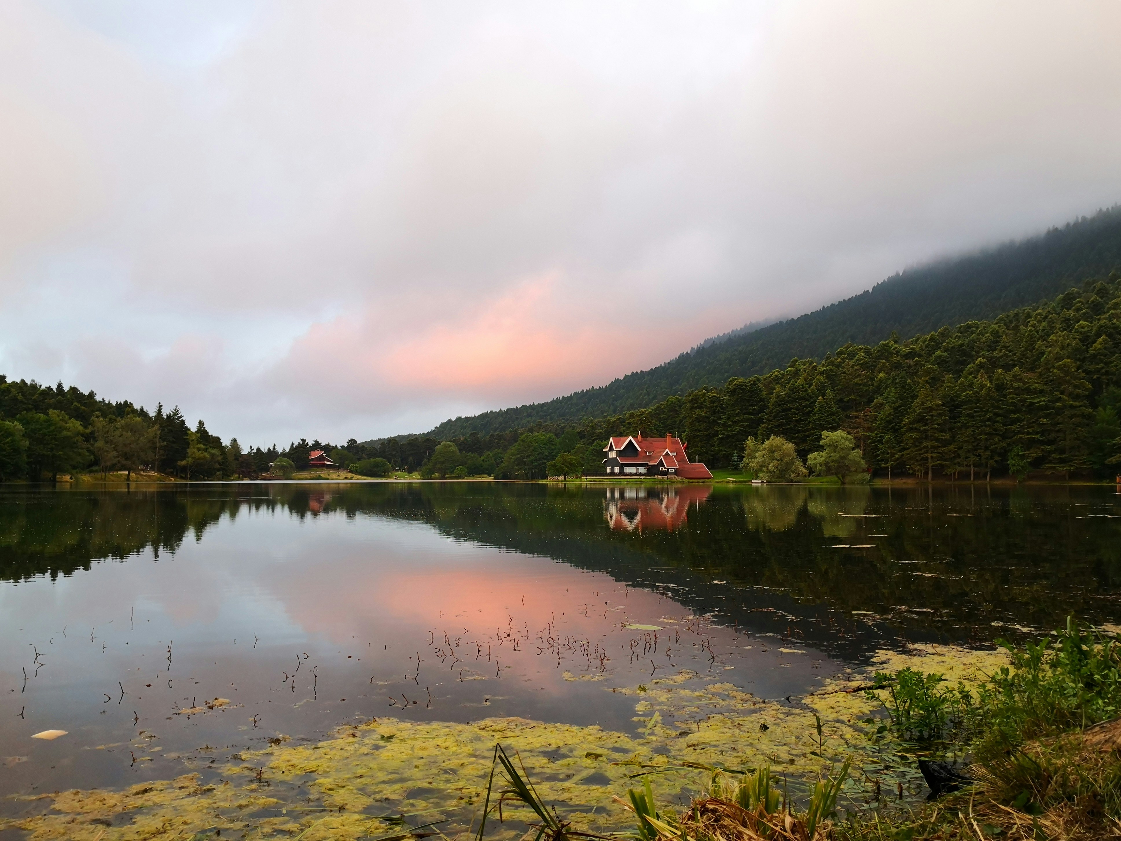 Calm lake reflecting a colorful sunset sky with distant houses and lush forested hills.
