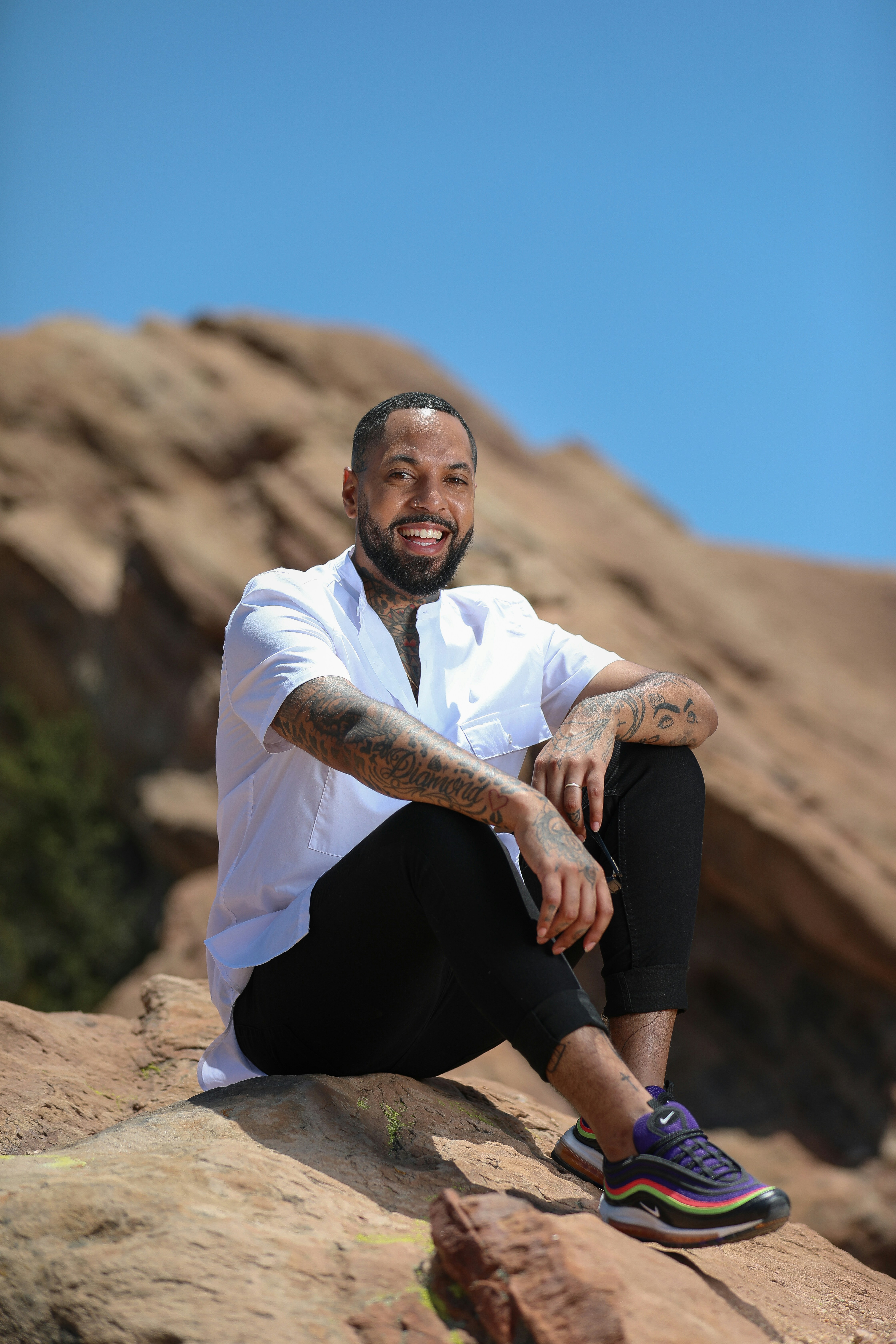 Man sitting on a rocky outcrop, smiling under a clear blue sky, showcasing a relaxed and joyful demeanor.
