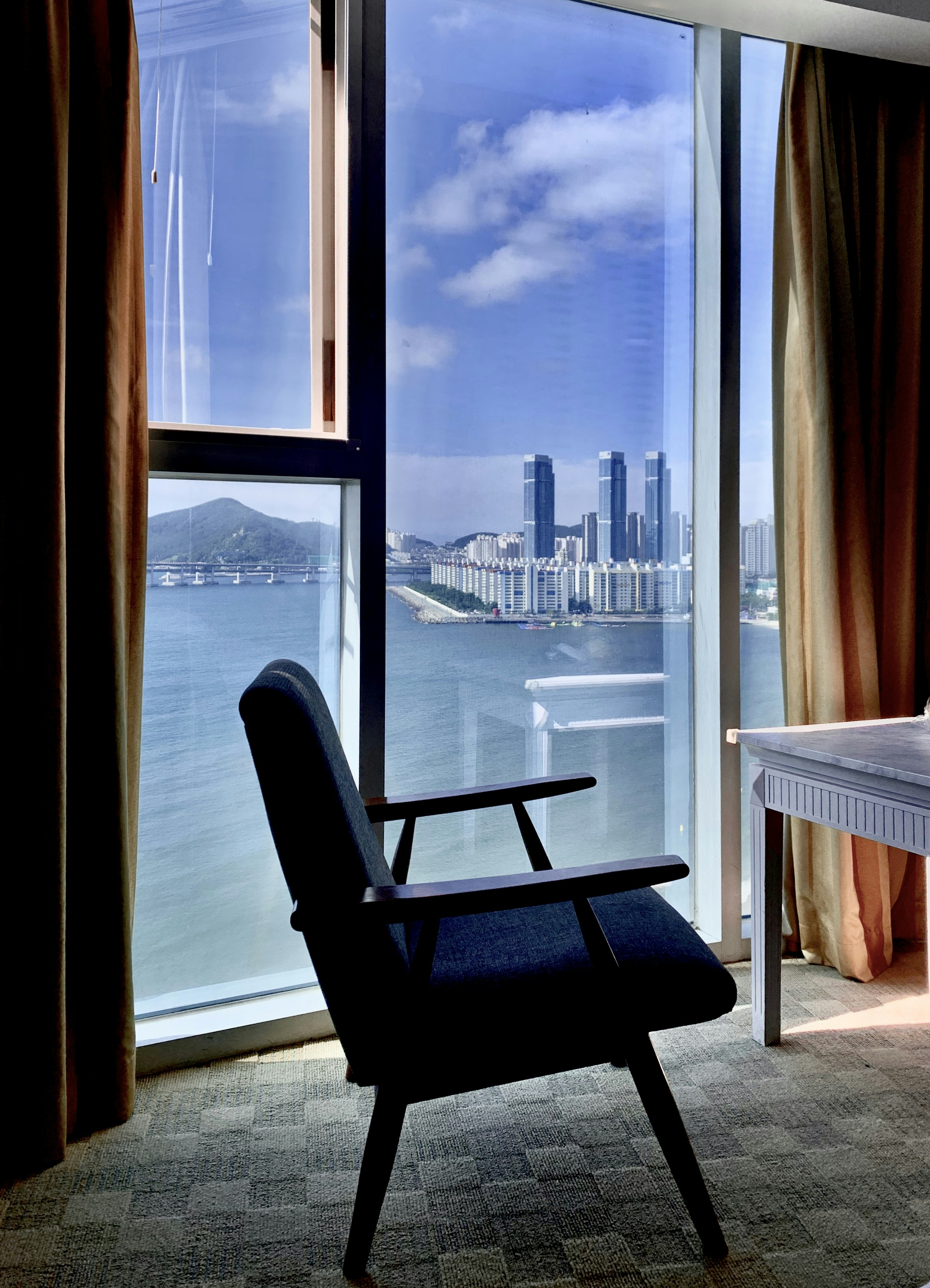 A cozy chair positioned by a large window, overlooking a serene waterfront view with modern skyscrapers and distant hills. The scene captures a blend of indoor comfort and outdoor beauty.