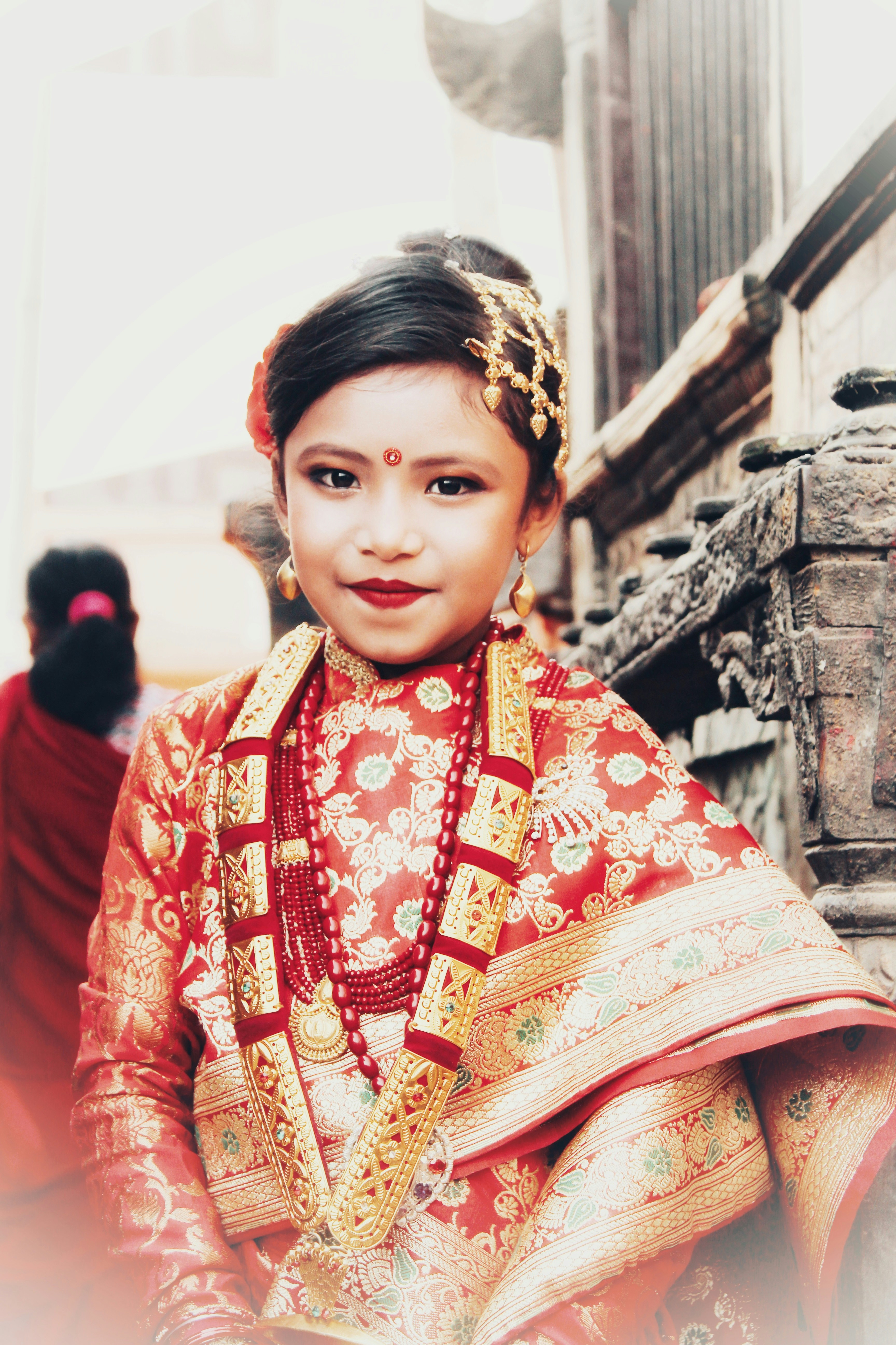 A young girl adorned in vibrant traditional attire, showcasing intricate jewelry and cultural significance. The backdrop hints at a historic setting.