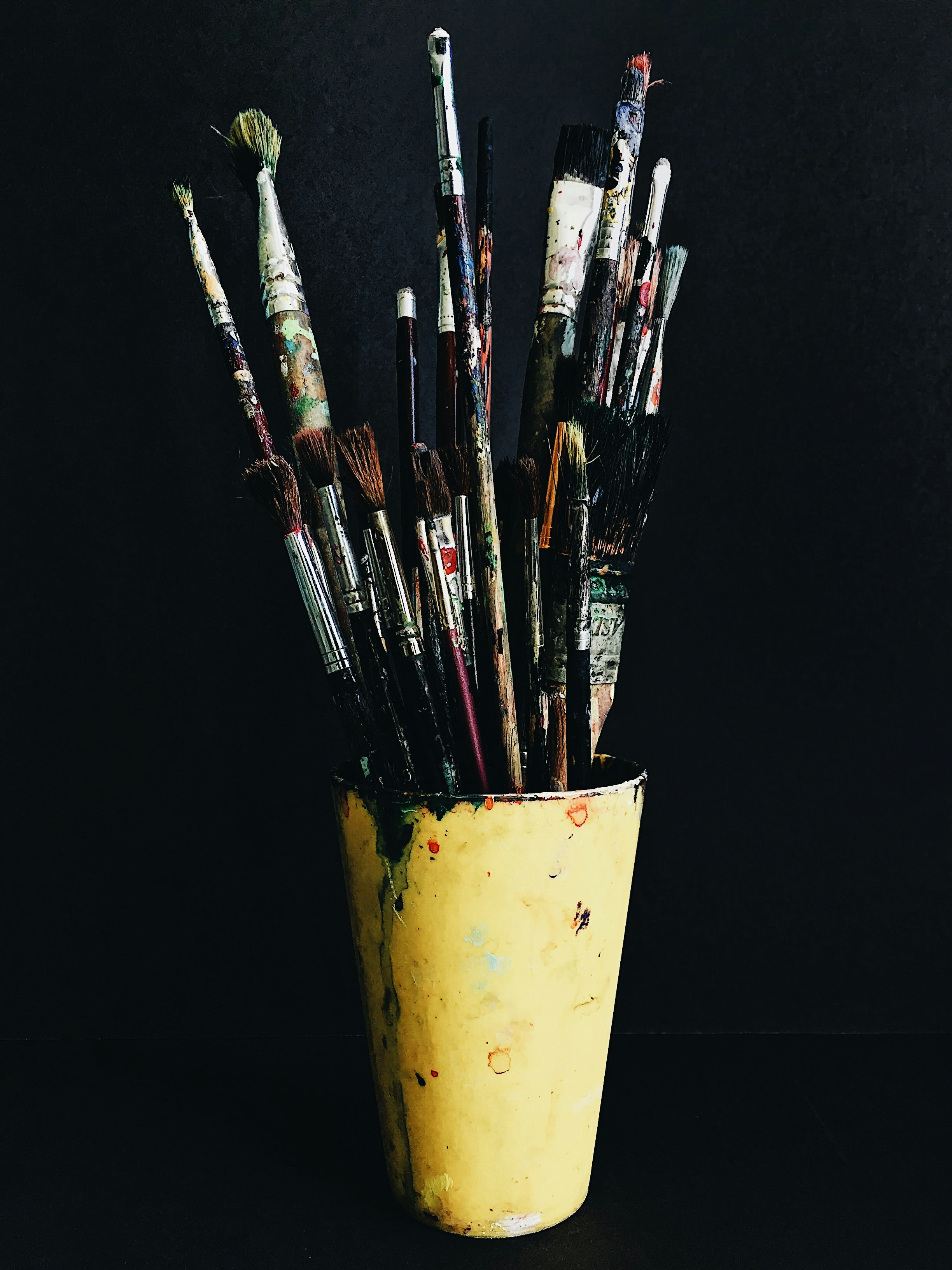 Collection of paintbrushes in a worn yellow container, showcasing vibrant colors and textures against a dark backdrop.
