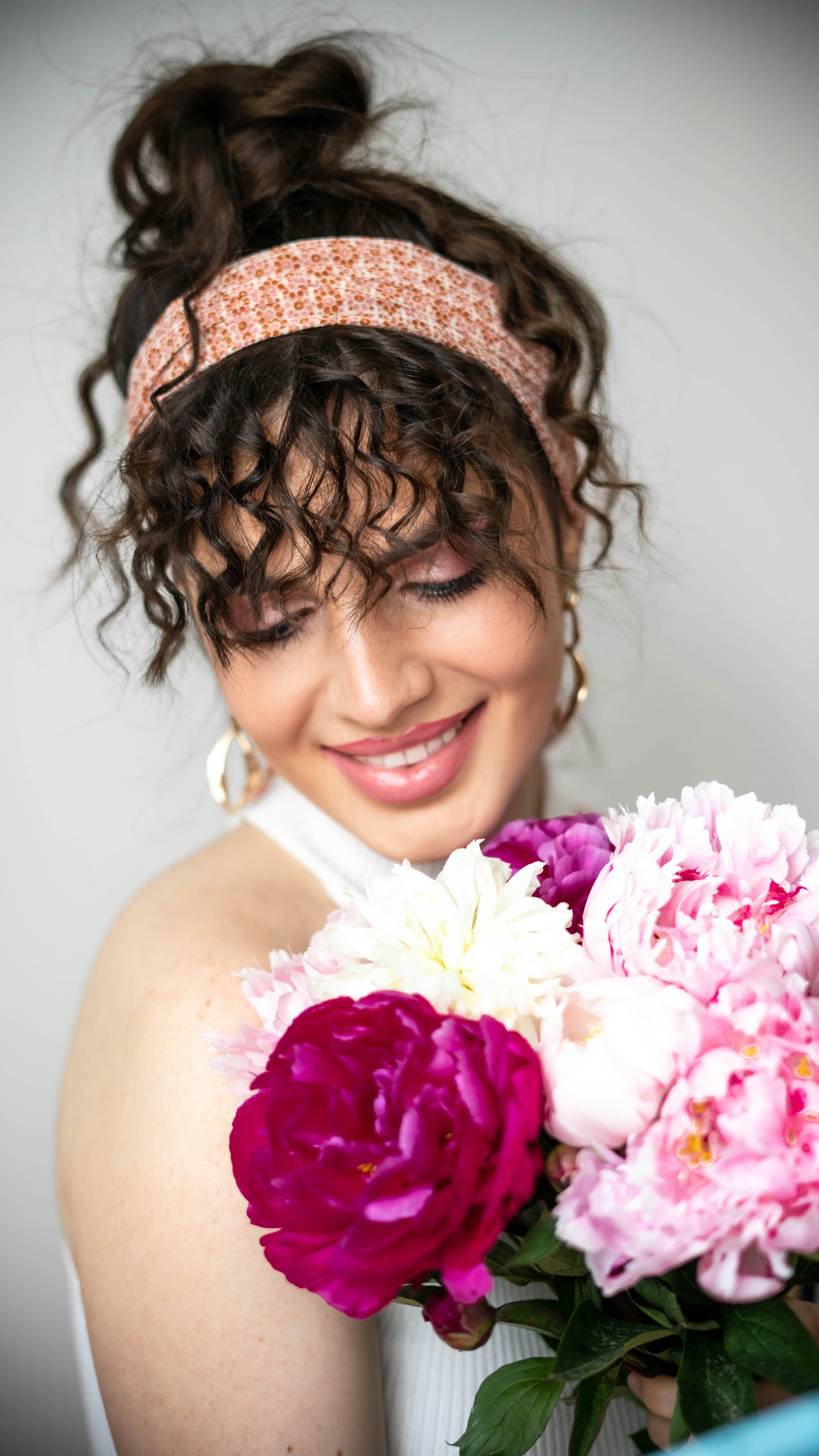 Woman with curly hair holding a vibrant bouquet of peonies, radiating warmth and happiness against a soft backdrop.