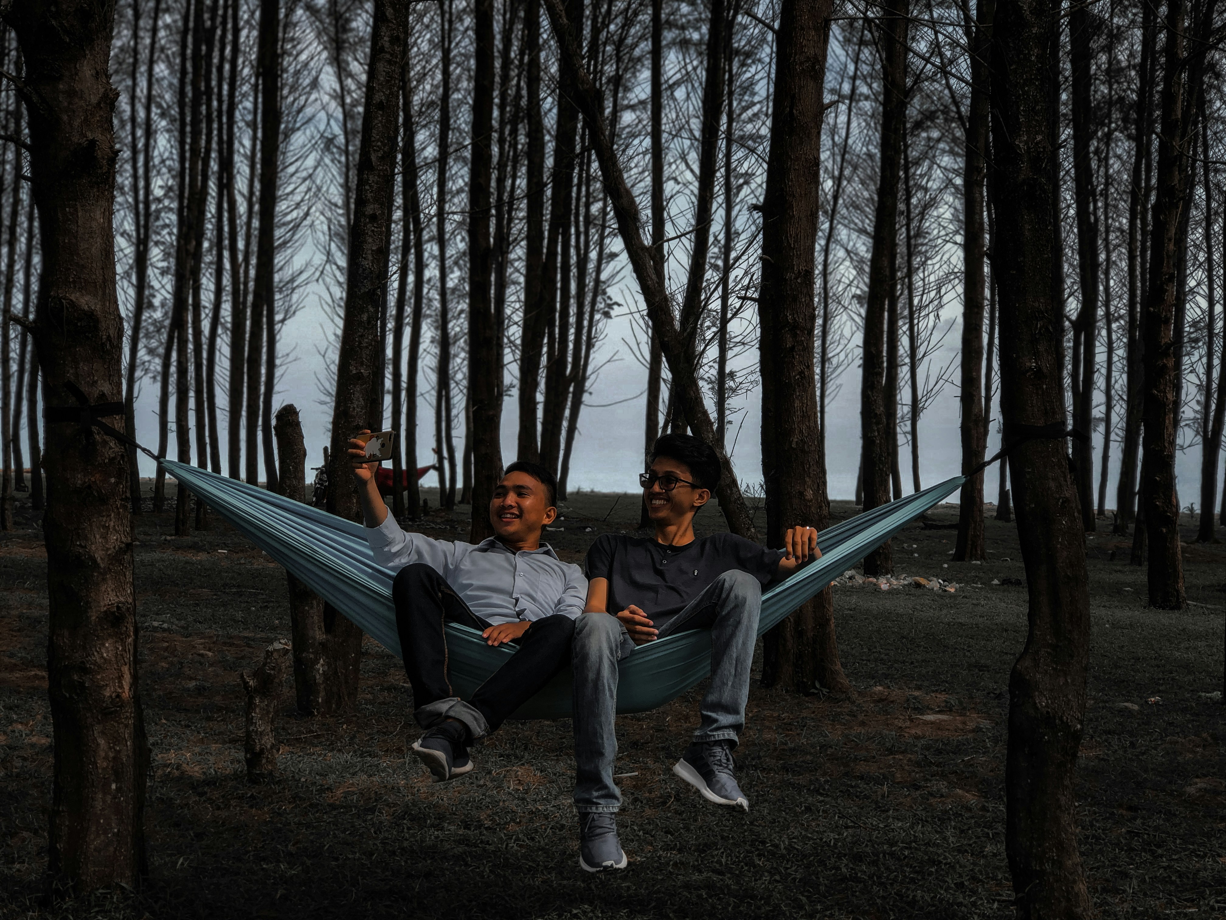 Two people relaxing on a hammock between tall trees in a dimly lit forest.