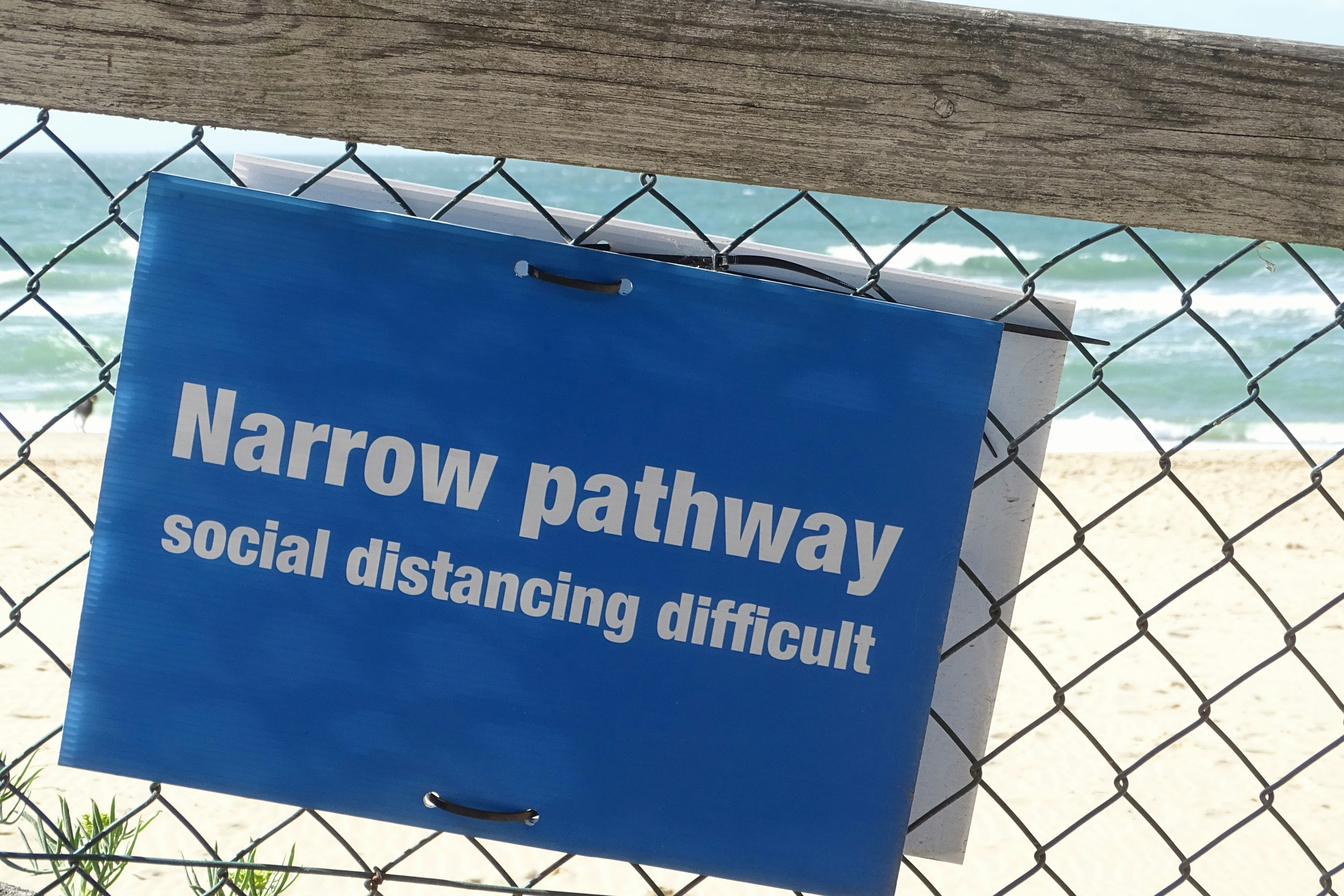 Sign warning about narrow pathway and social distancing challenges near a beach. The text is clearly visible against a blue background.