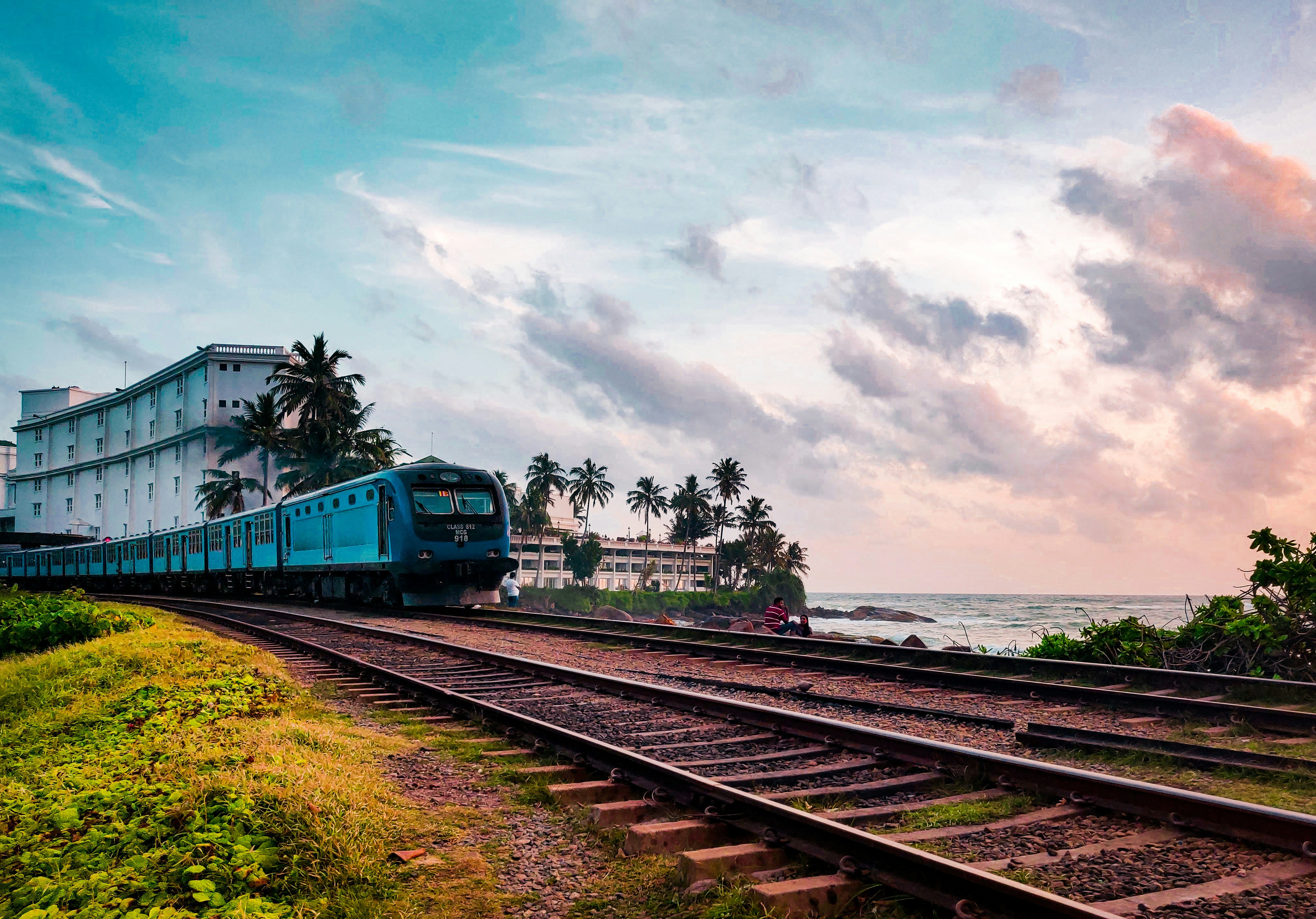 A blue train glides along the railway beside a tranquil coastline, with palm trees and a hotel in the background. The sky is painted with soft pastel hues of sunset.