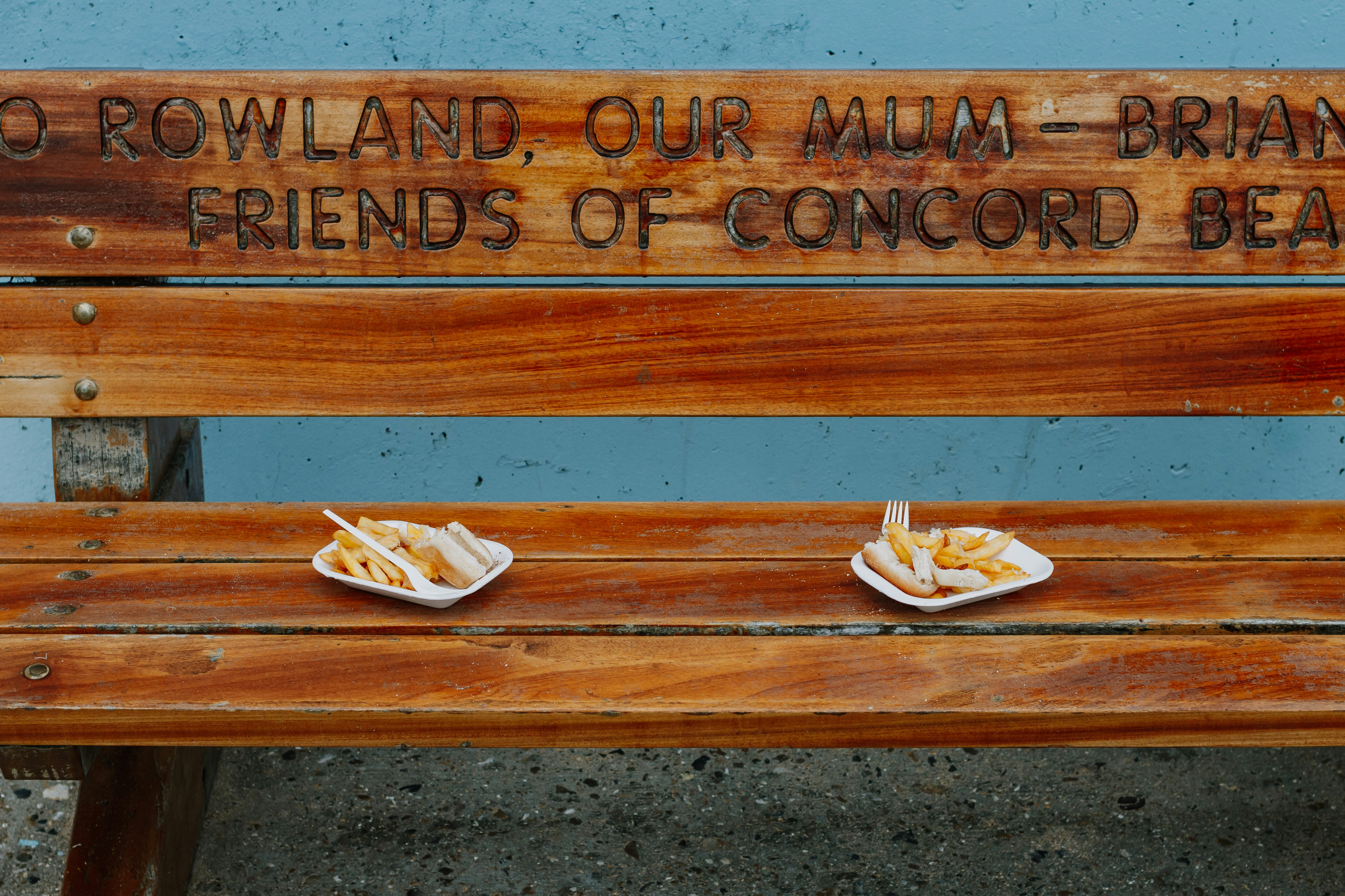 Two plates of fries sit on a weathered wooden bench engraved with a heartfelt dedication. The scene captures a moment of leisure and community spirit.