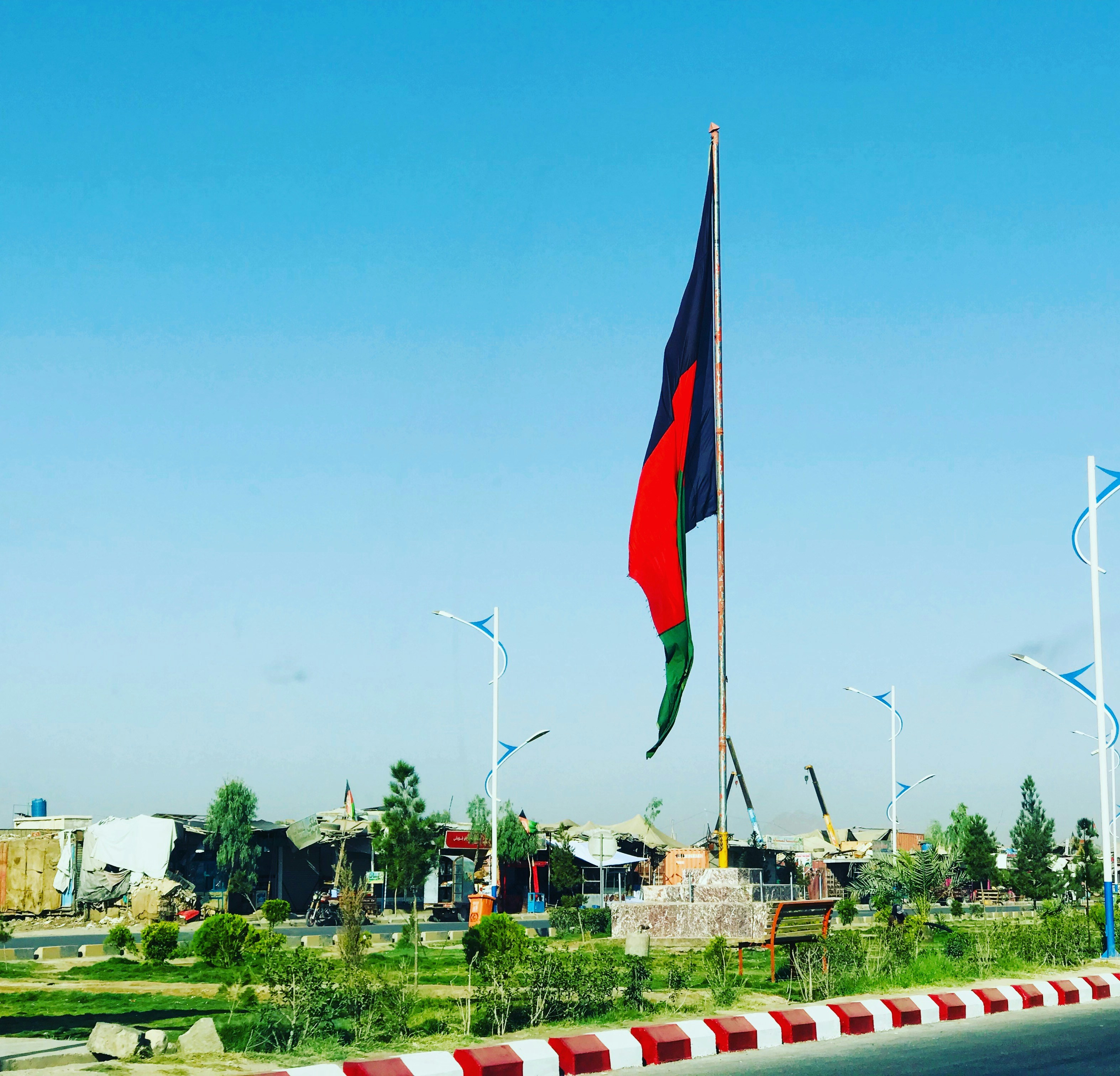 A large Afghan flag waves proudly over a bustling area, surrounded by greenery and modern streetlights. The scene captures a blend of tradition and contemporary life.