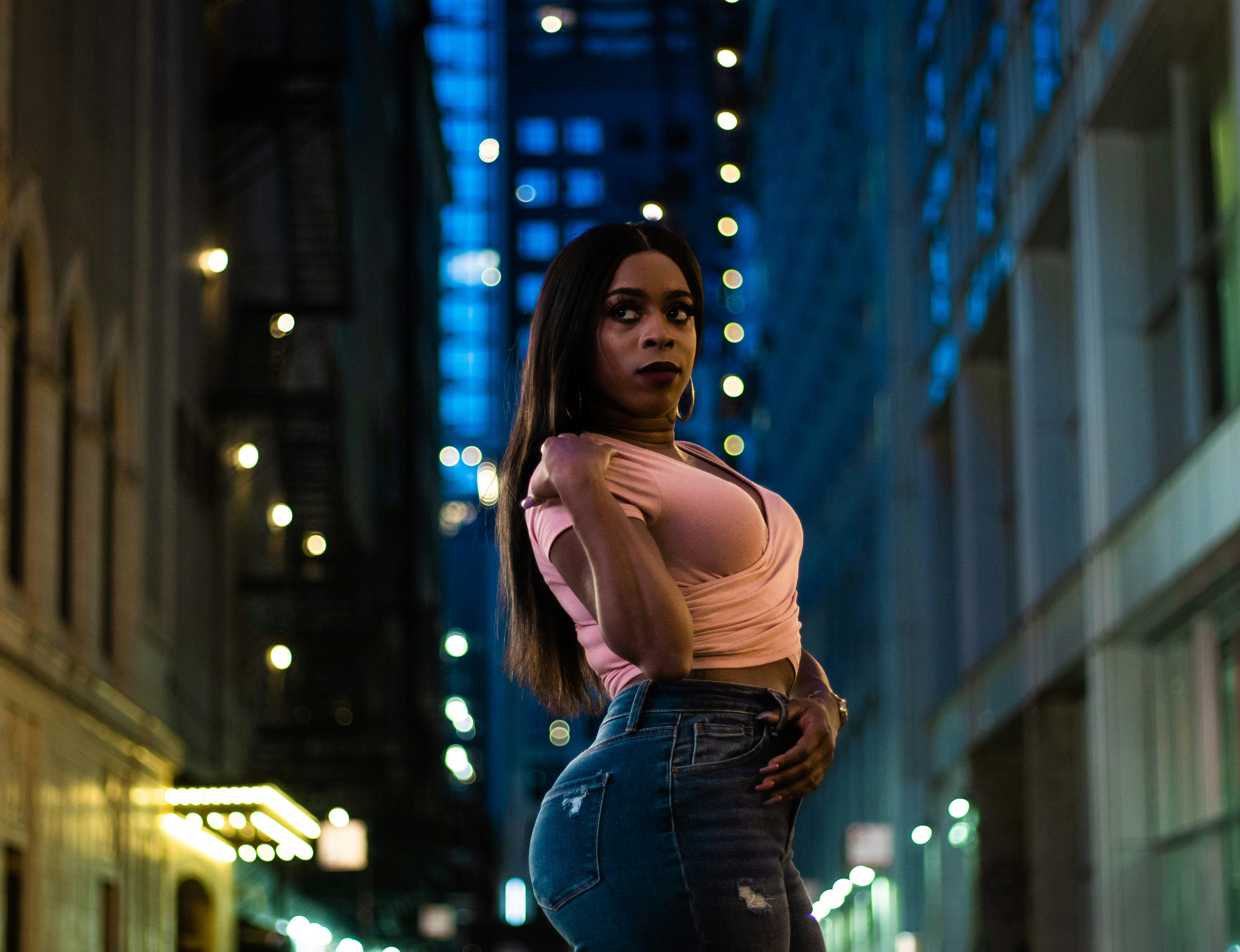 A confident figure poses in a city alley, illuminated by soft blue and warm lights, showcasing a blend of urban style and nighttime allure.