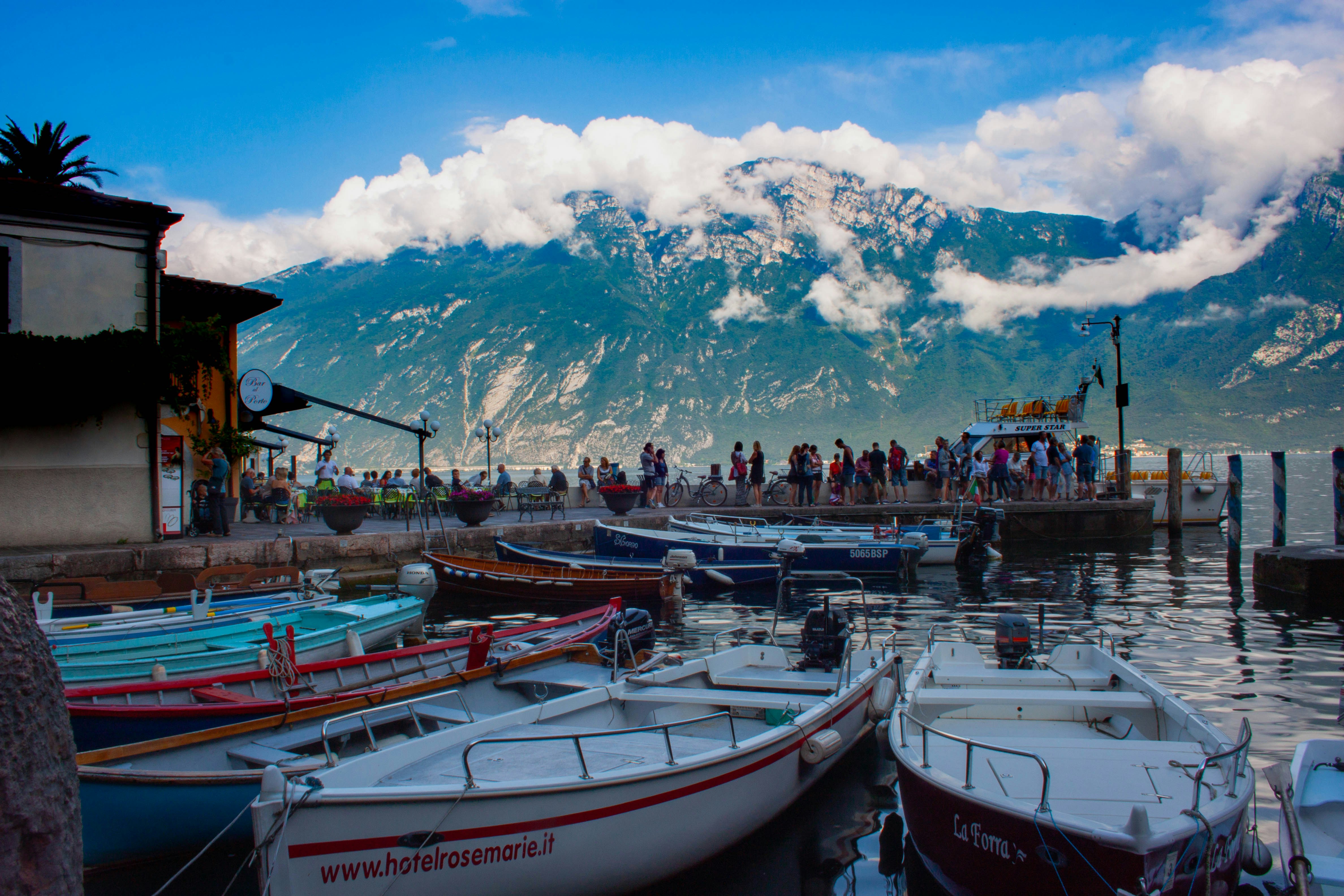 Colorful boats lined up at a serene lakeside harbor with mountains and clouds in the background.