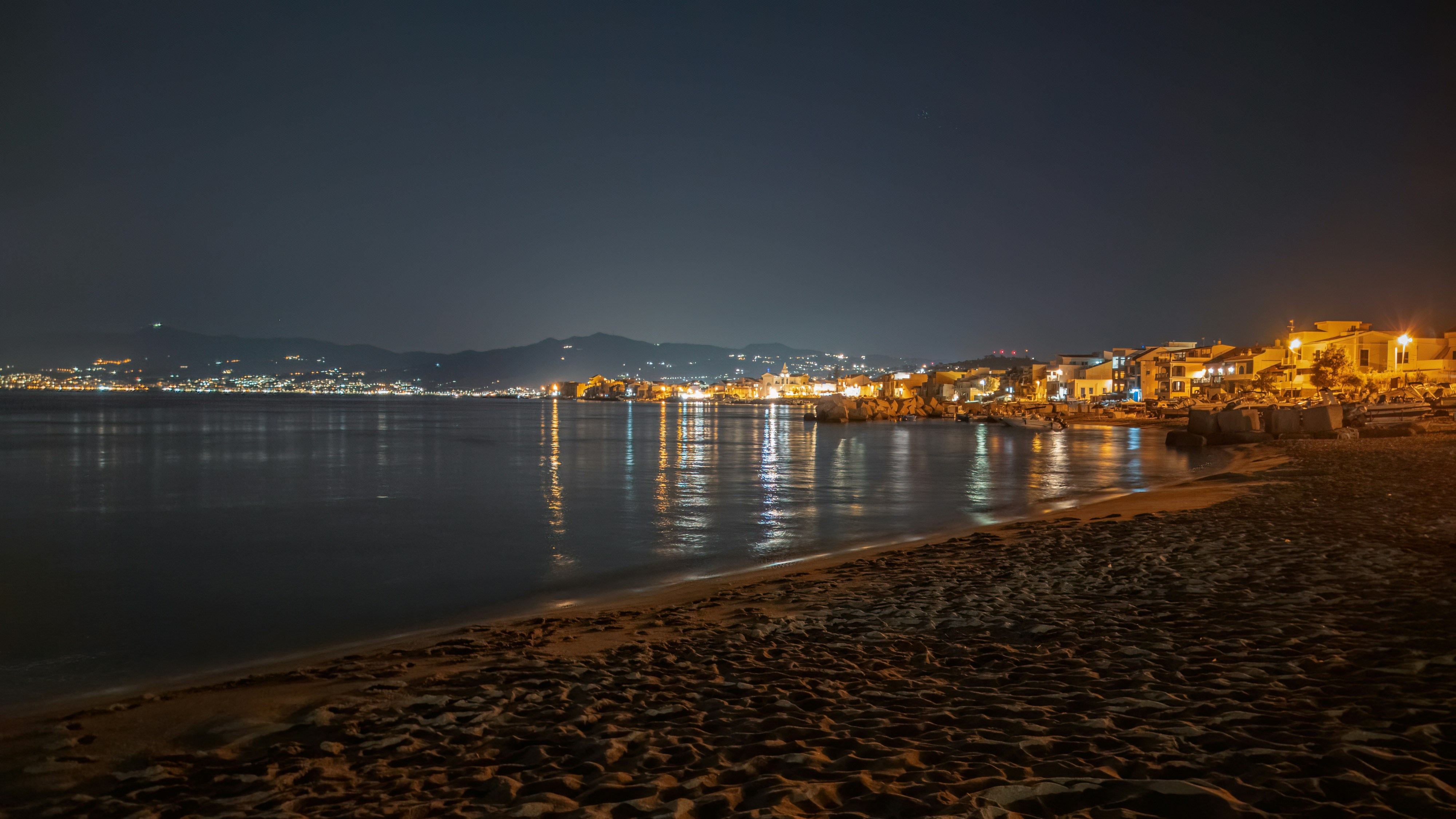 Coastal town illuminated by warm lights along the shoreline at night, with gentle waves reflecting the vibrant colors. 