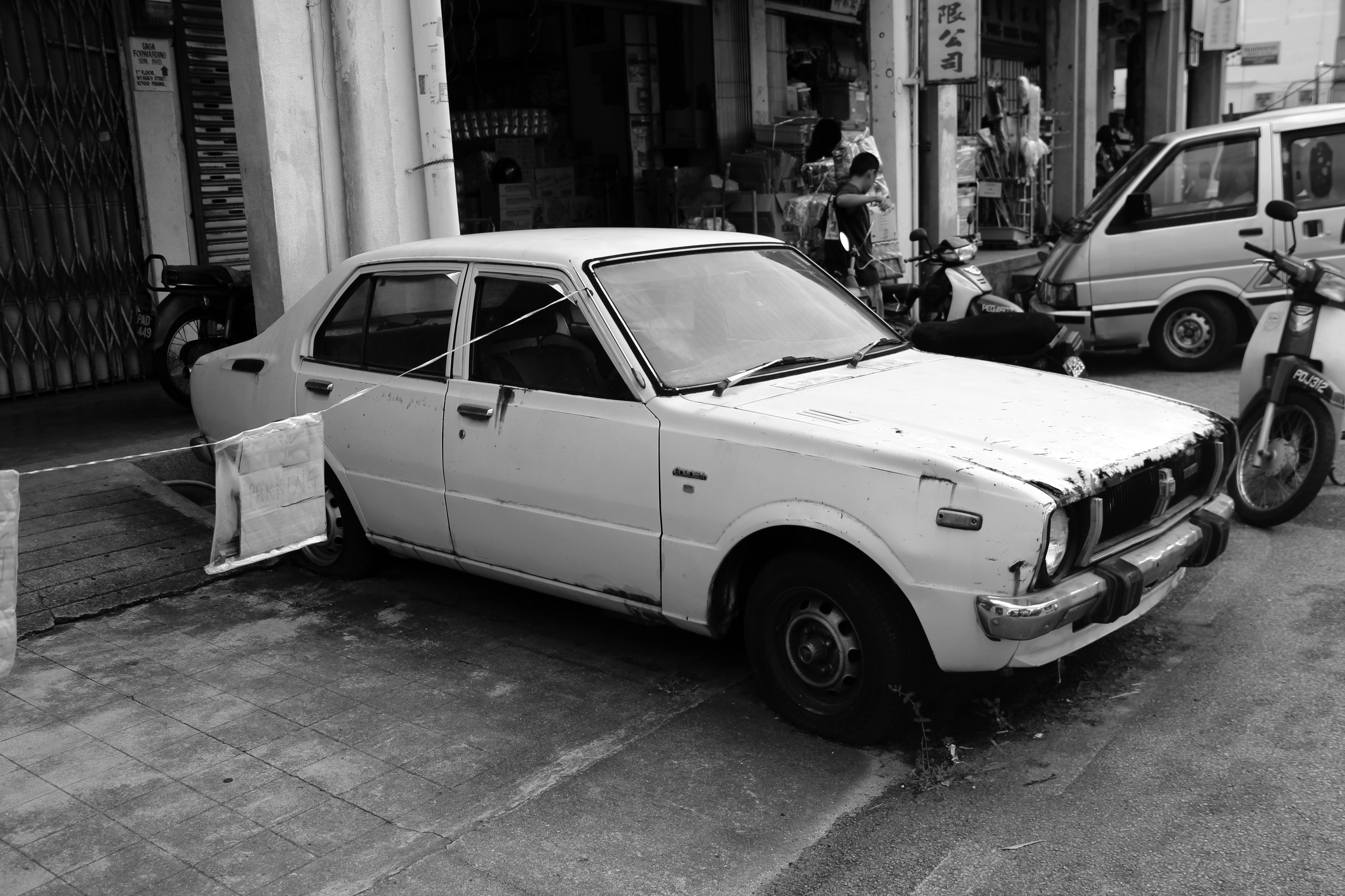 An aged white sedan parked on a city street, surrounded by bustling shops and motorcycles, showcasing urban decay and nostalgia.