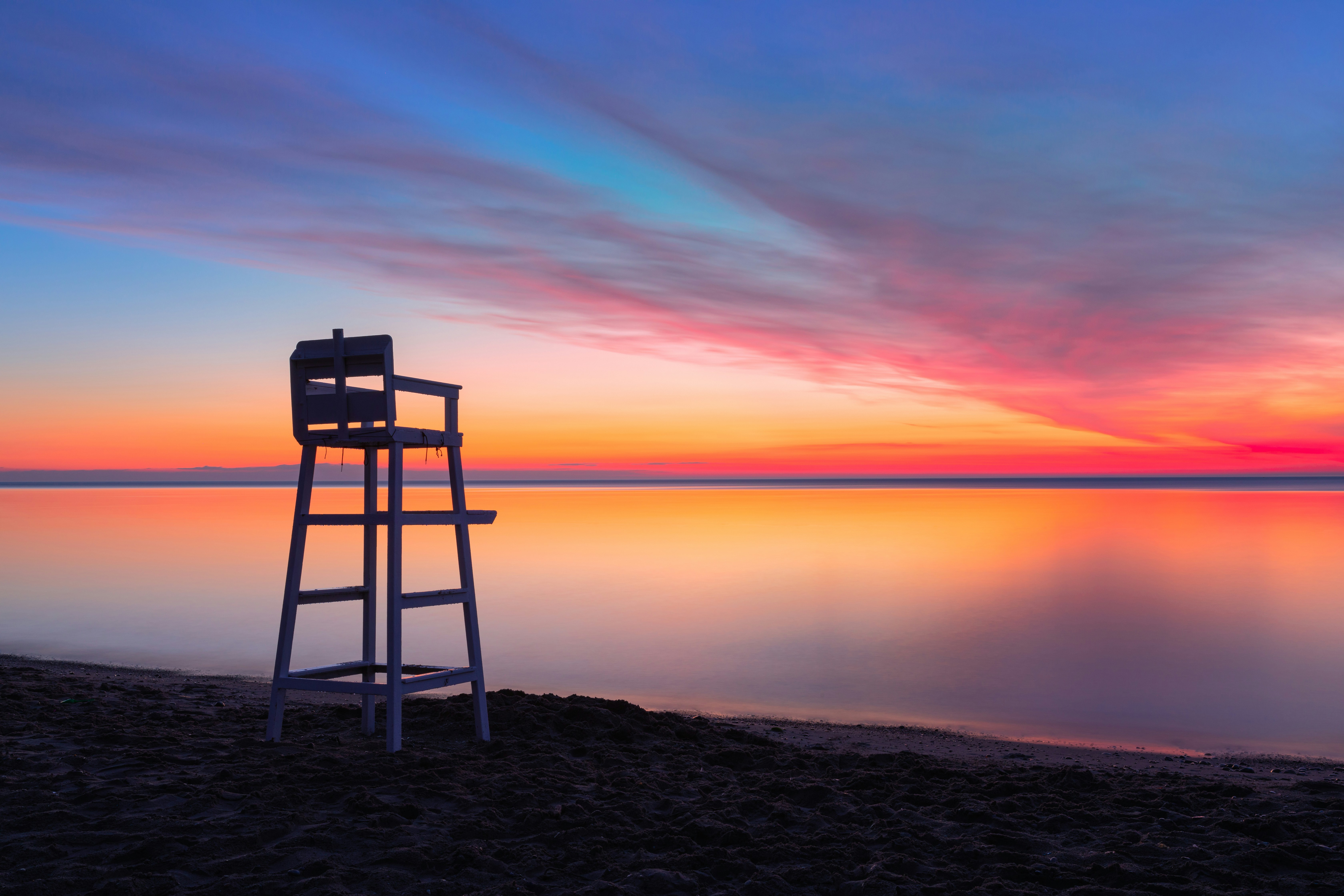 Solitary lifeguard chair silhouetted against a vibrant, multicolored sunset over calm waters.