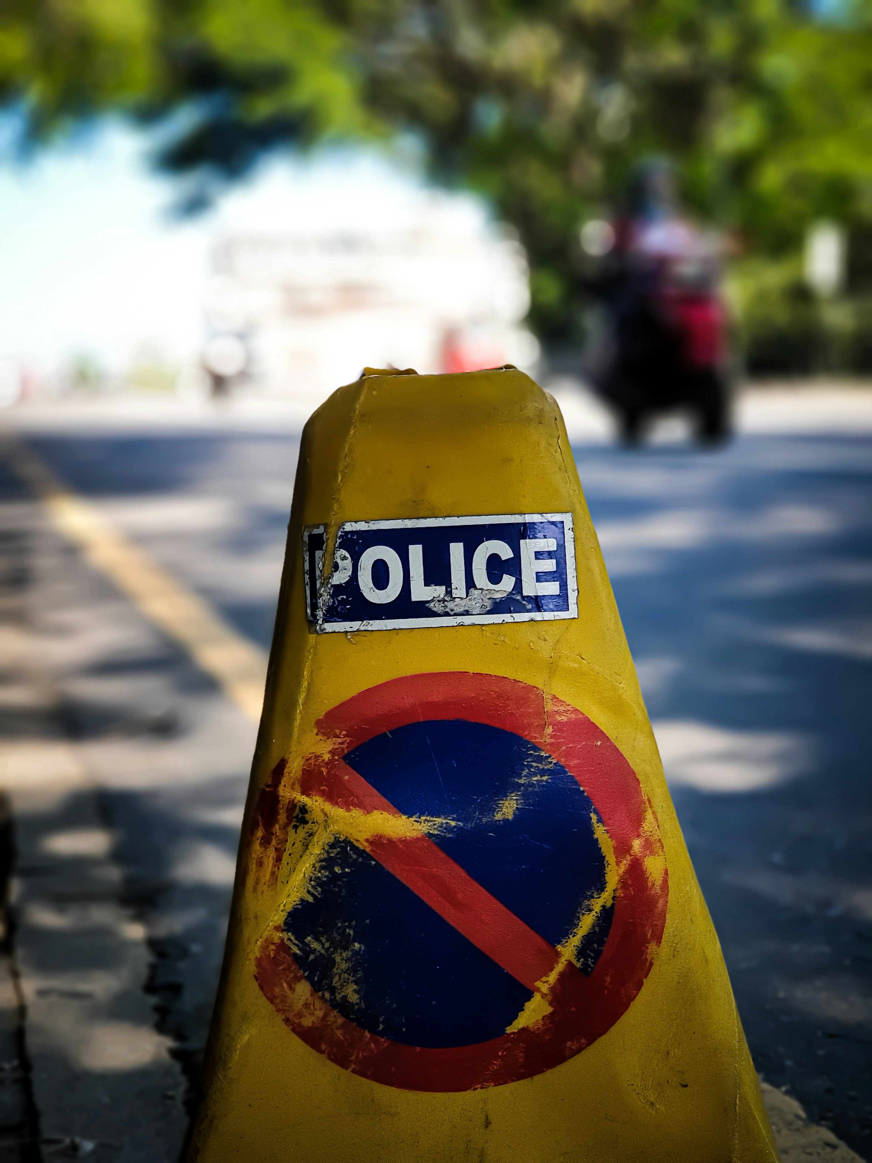 Yellow traffic cone with 'POLICE' label and a no-parking symbol, positioned on a road. The background features blurred vehicles and greenery.