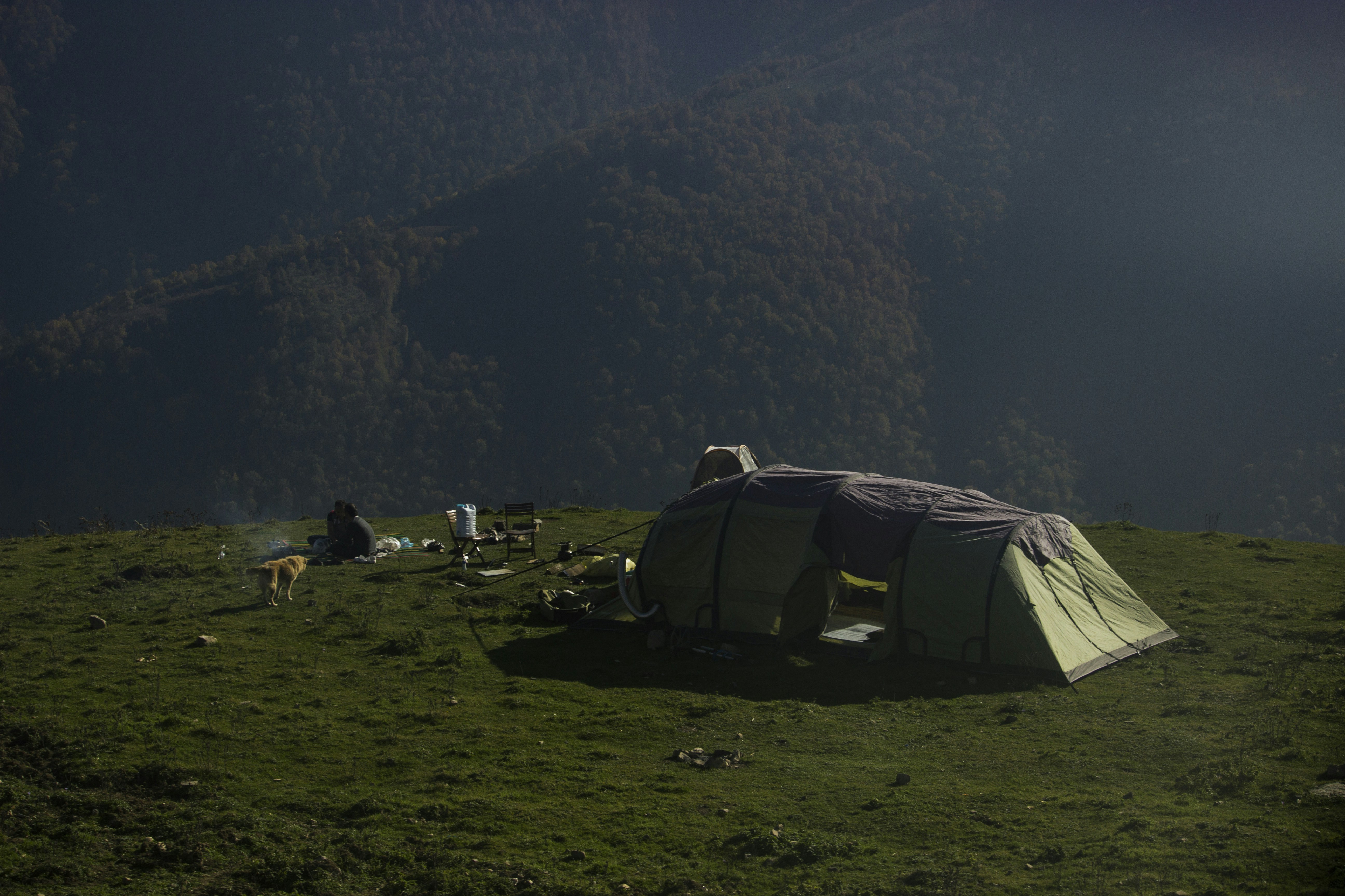 A green tent set against a mountainous backdrop, with a person and a dog enjoying a peaceful moment outdoors. Smoke from a campfire rises gently in the air.