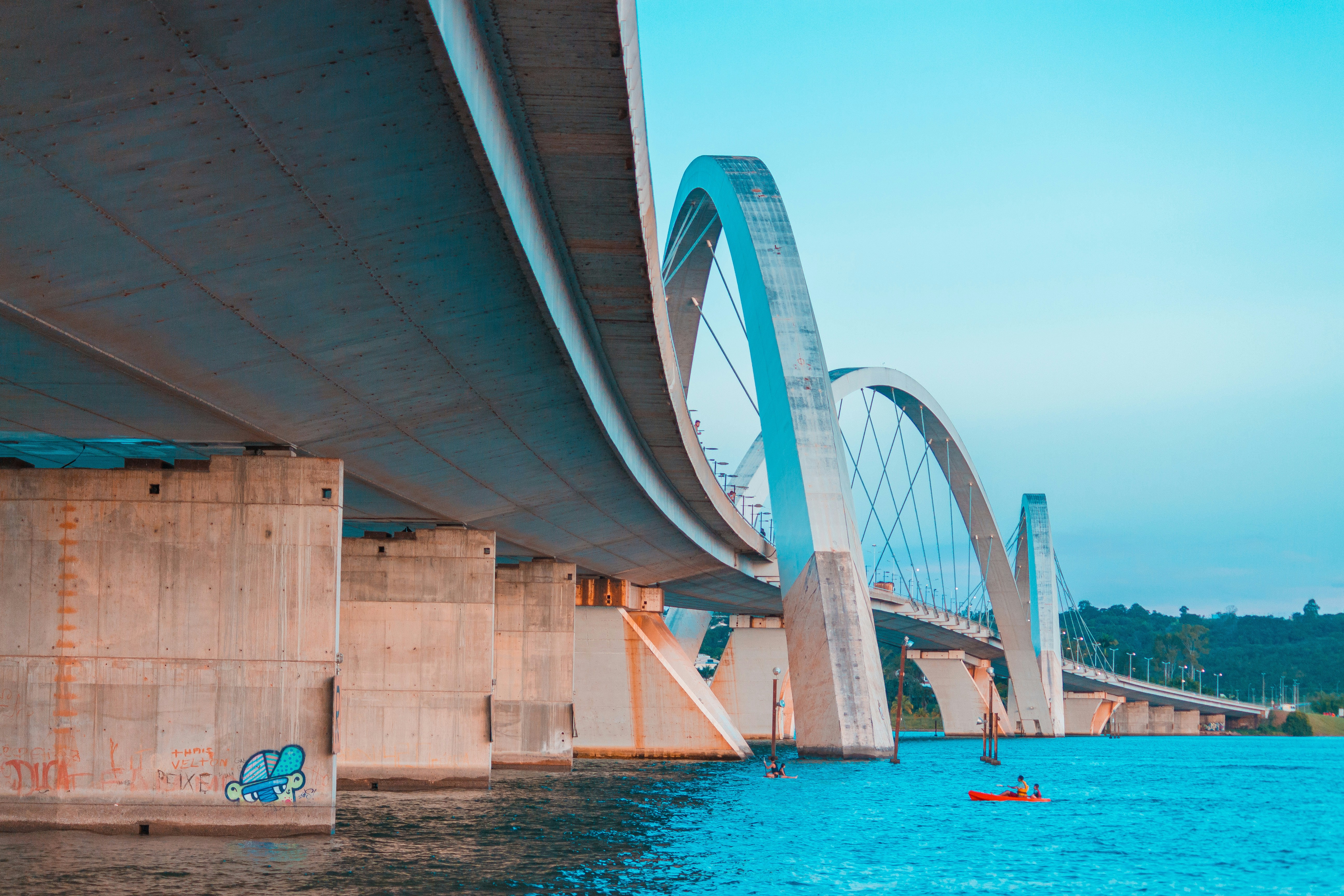 Sleek architectural bridge structure reflected in calm water, with a kayak navigating beneath. Graffiti art adds an urban touch to the concrete pillars.