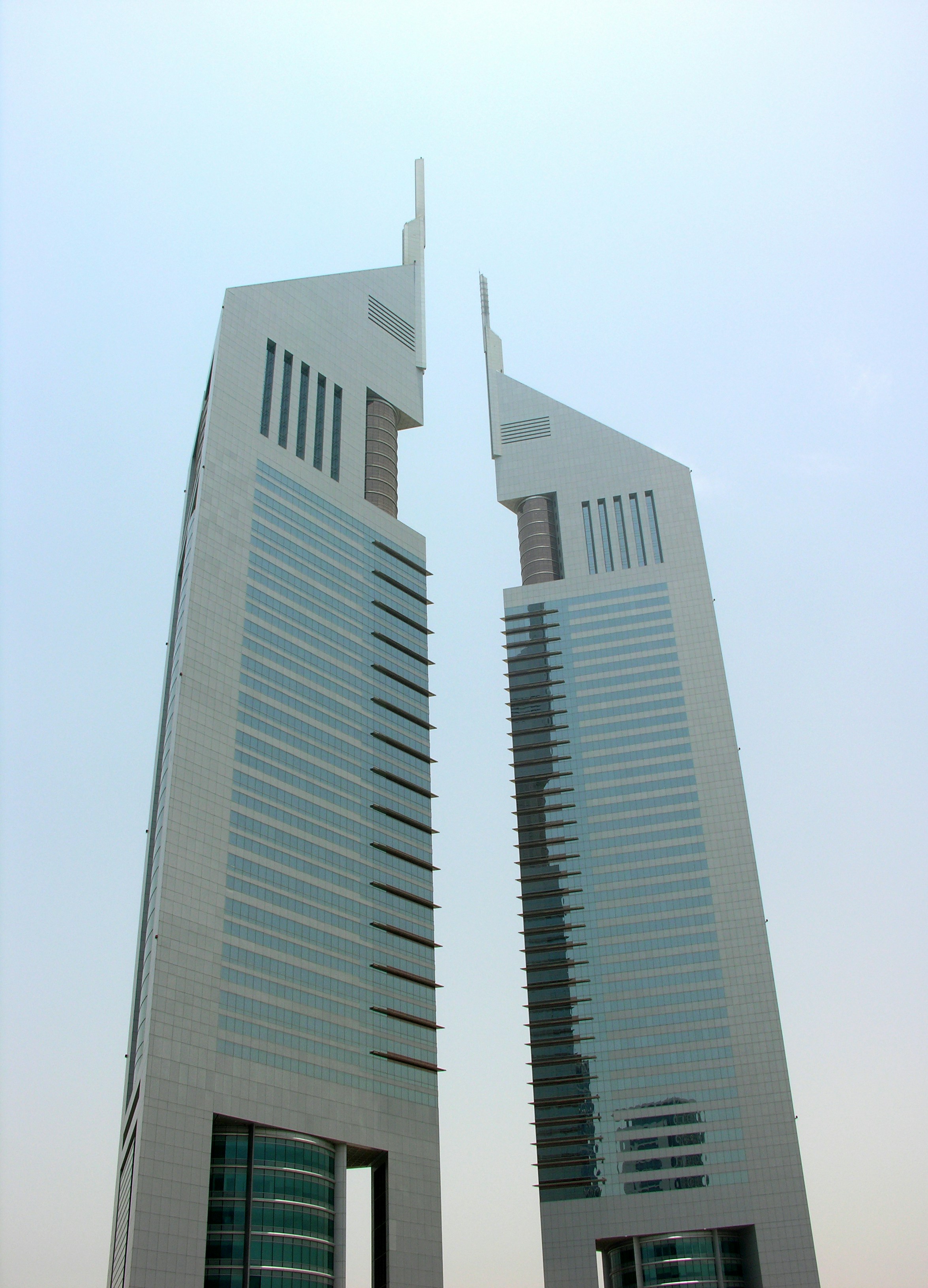 Two modern skyscrapers with sleek glass facades and distinctive architectural features against a pale sky.