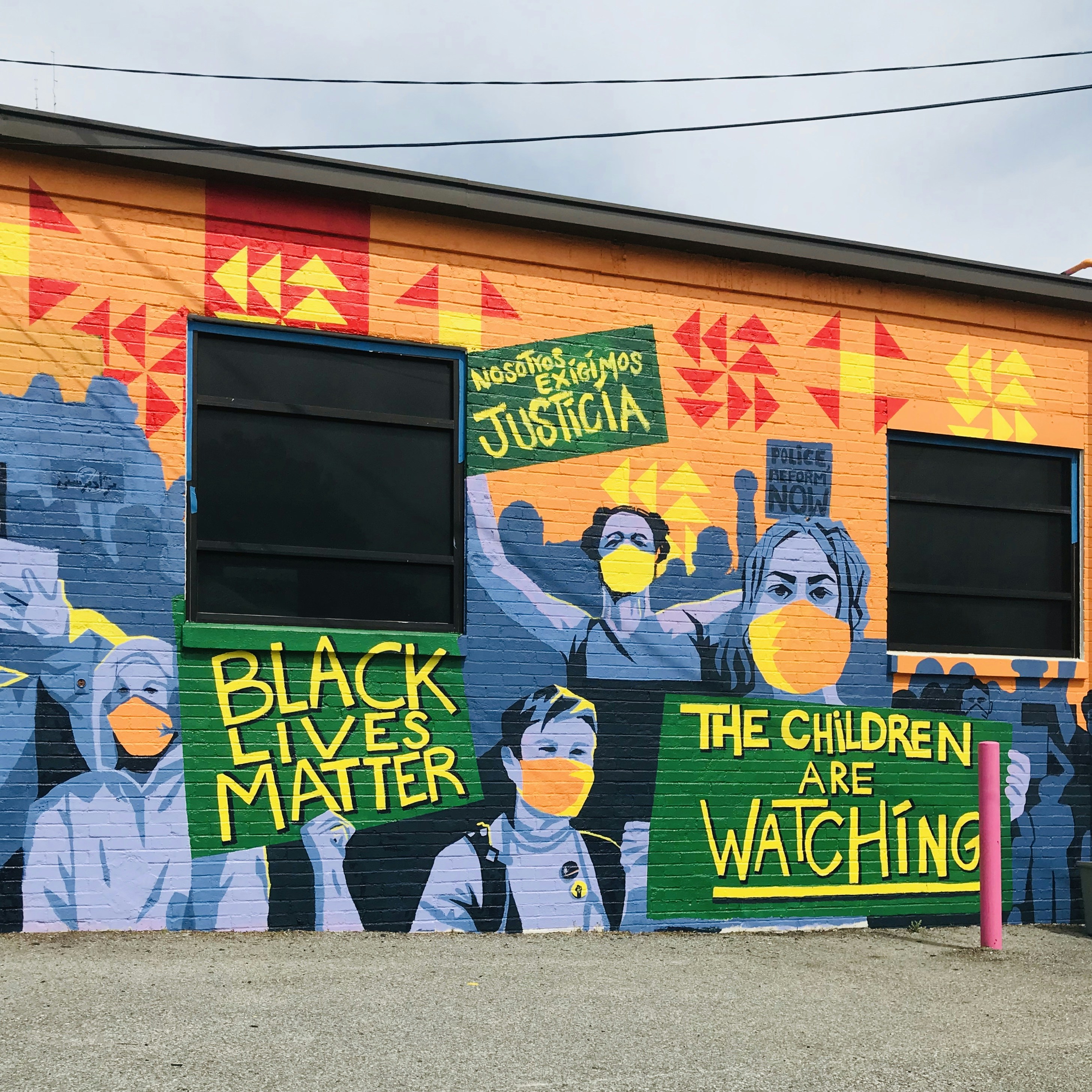Vibrant mural featuring diverse figures wearing masks, advocating for social justice with bold slogans. The artwork emphasizes community and awareness.