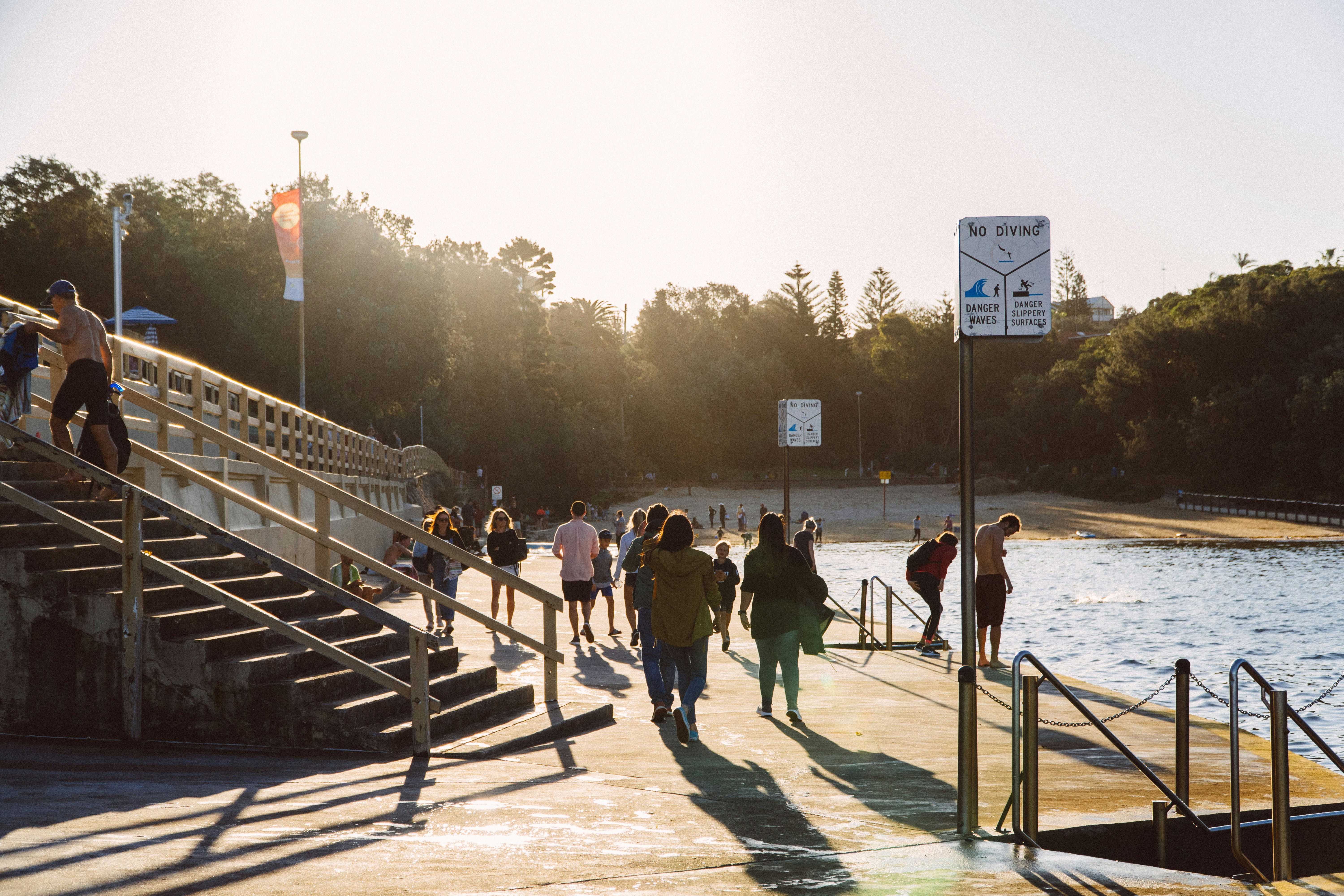 People enjoying a leisurely walk along a waterfront promenade as the sun sets, casting long shadows on the pavement.