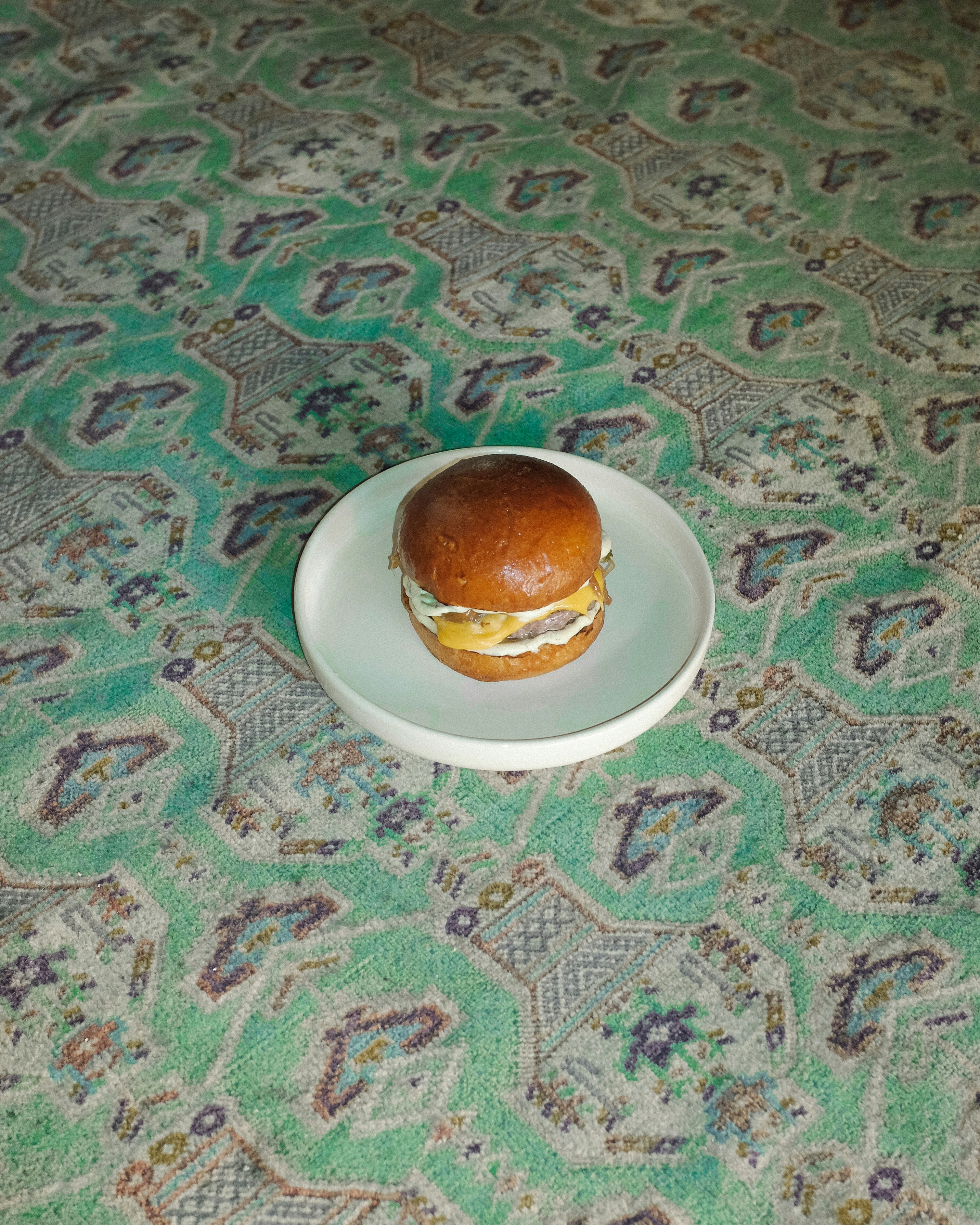 A freshly made burger resting on a white plate, set against an ornate green patterned carpet.