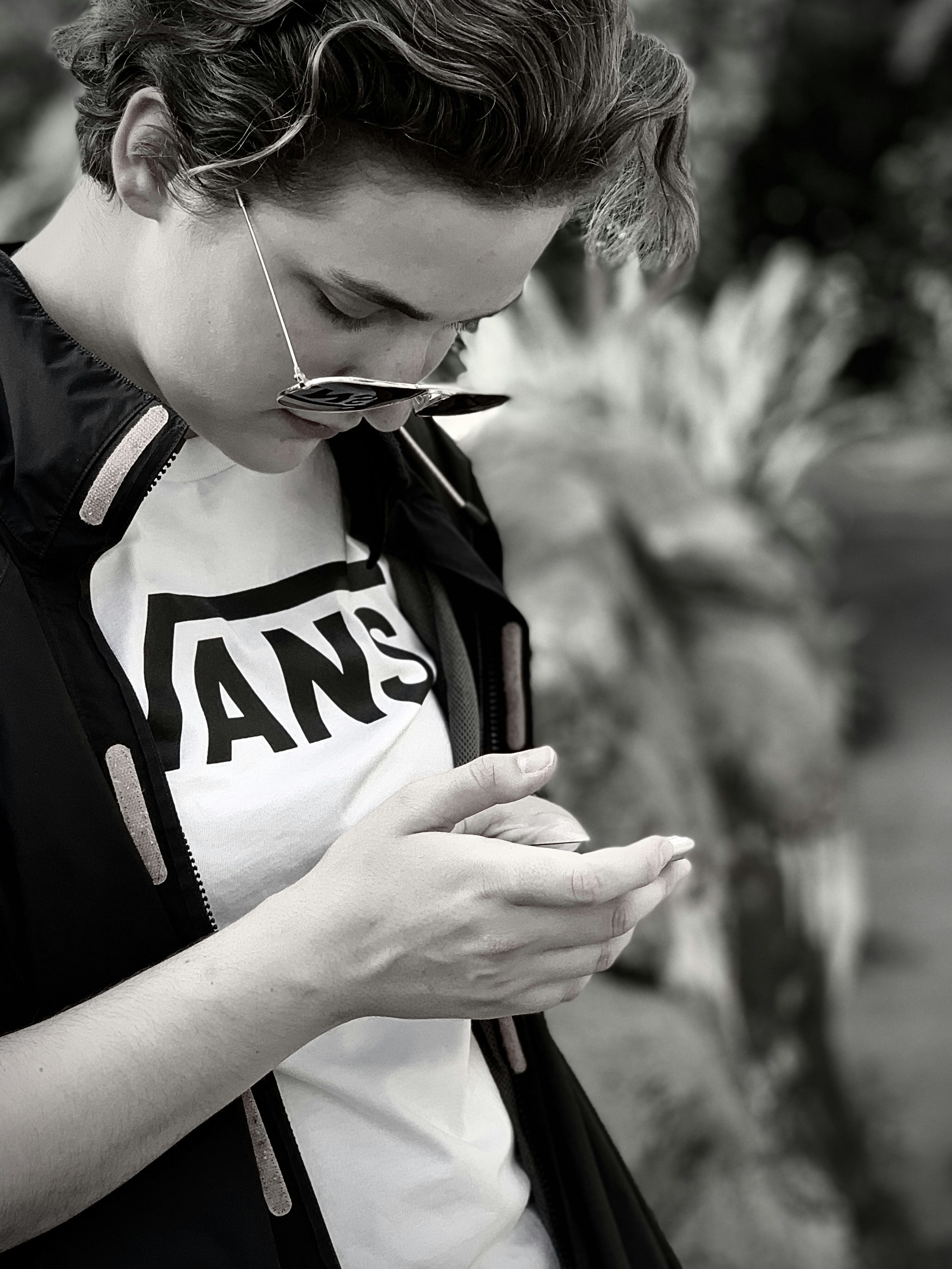 Young person in a black jacket and Vans t-shirt focused on a smartphone, with sunglasses resting on their nose. Background features blurred greenery.