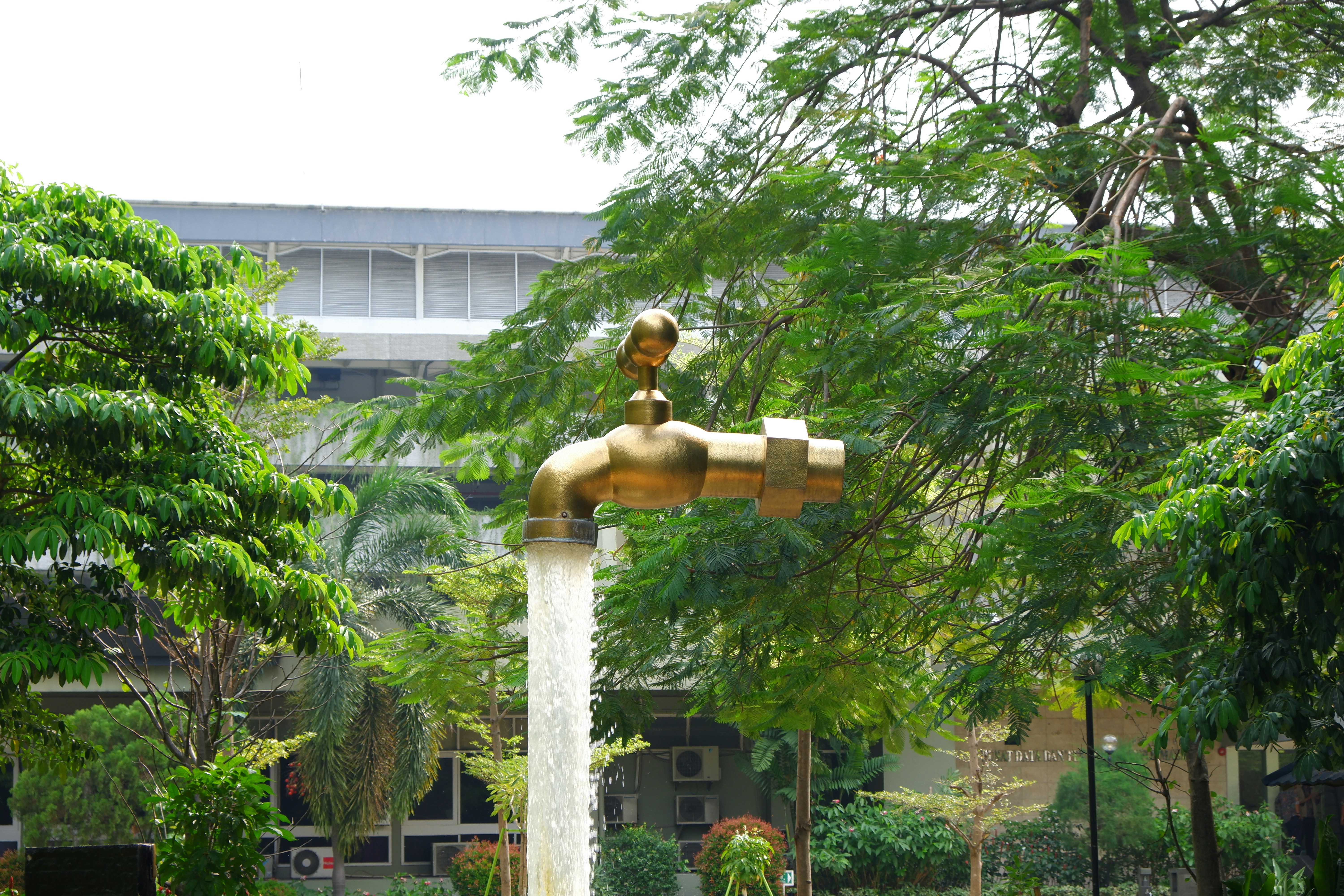 A brass faucet pouring water, surrounded by lush green trees in a vibrant outdoor setting.