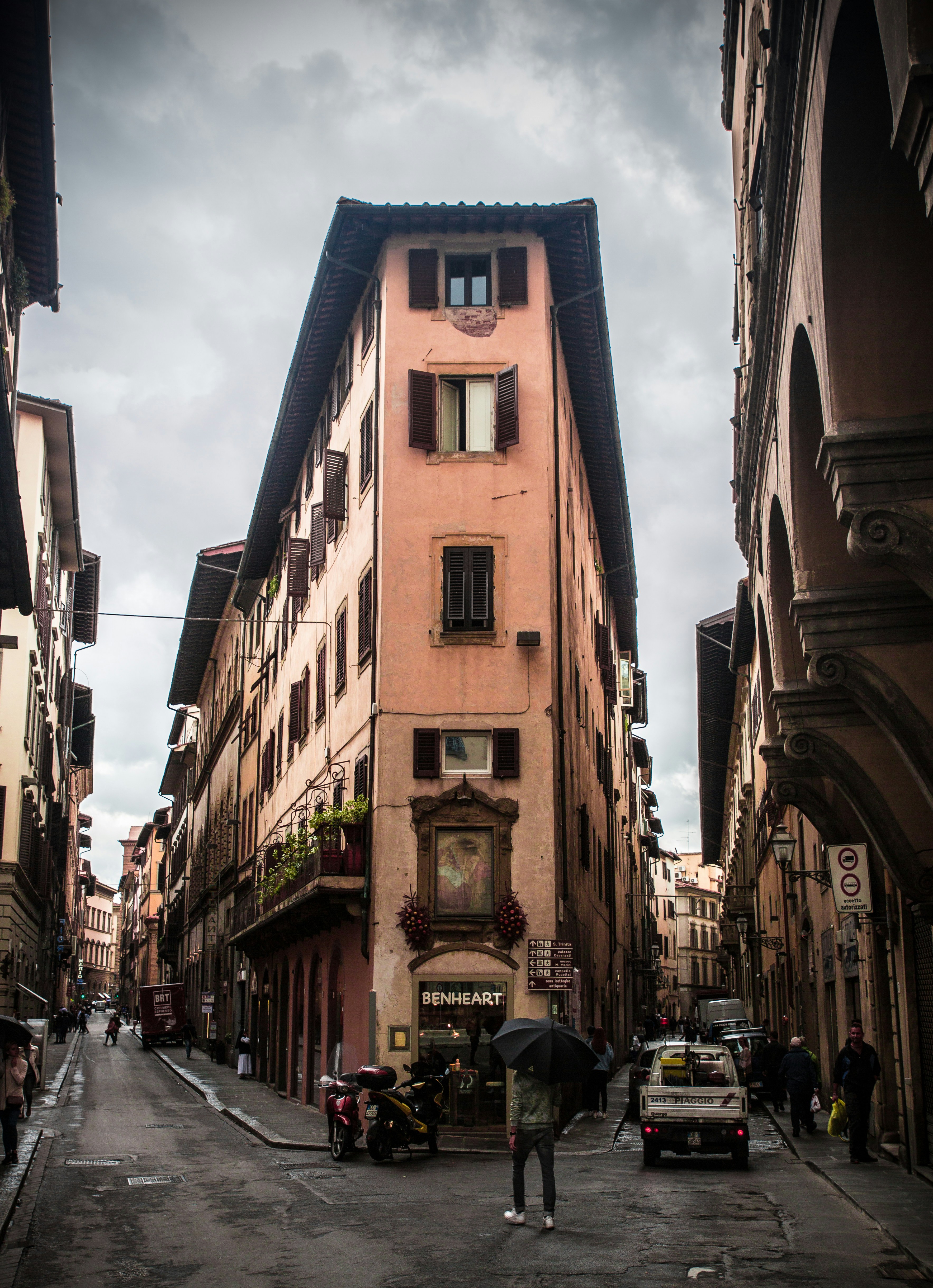 Narrow street in Florence showcasing a peach-colored building adorned with flowers and a vintage sign. A figure with an umbrella adds a touch of life to the scene.