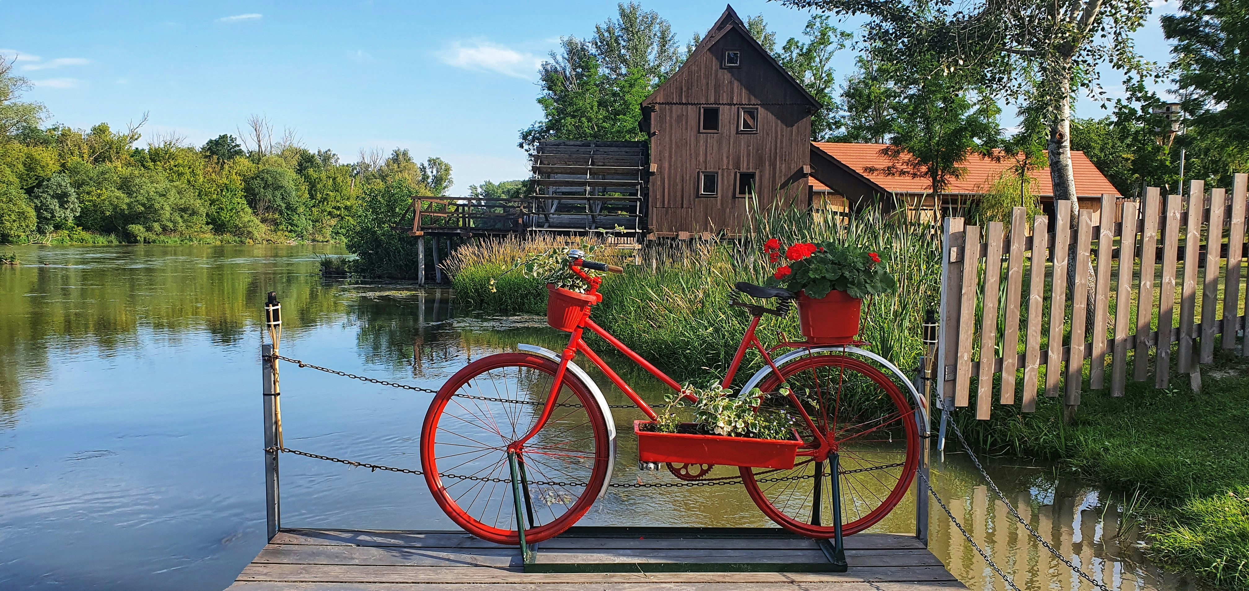 A vibrant red bicycle adorned with flowers stands on a dock beside a tranquil river, framed by lush greenery and rustic buildings in the background.