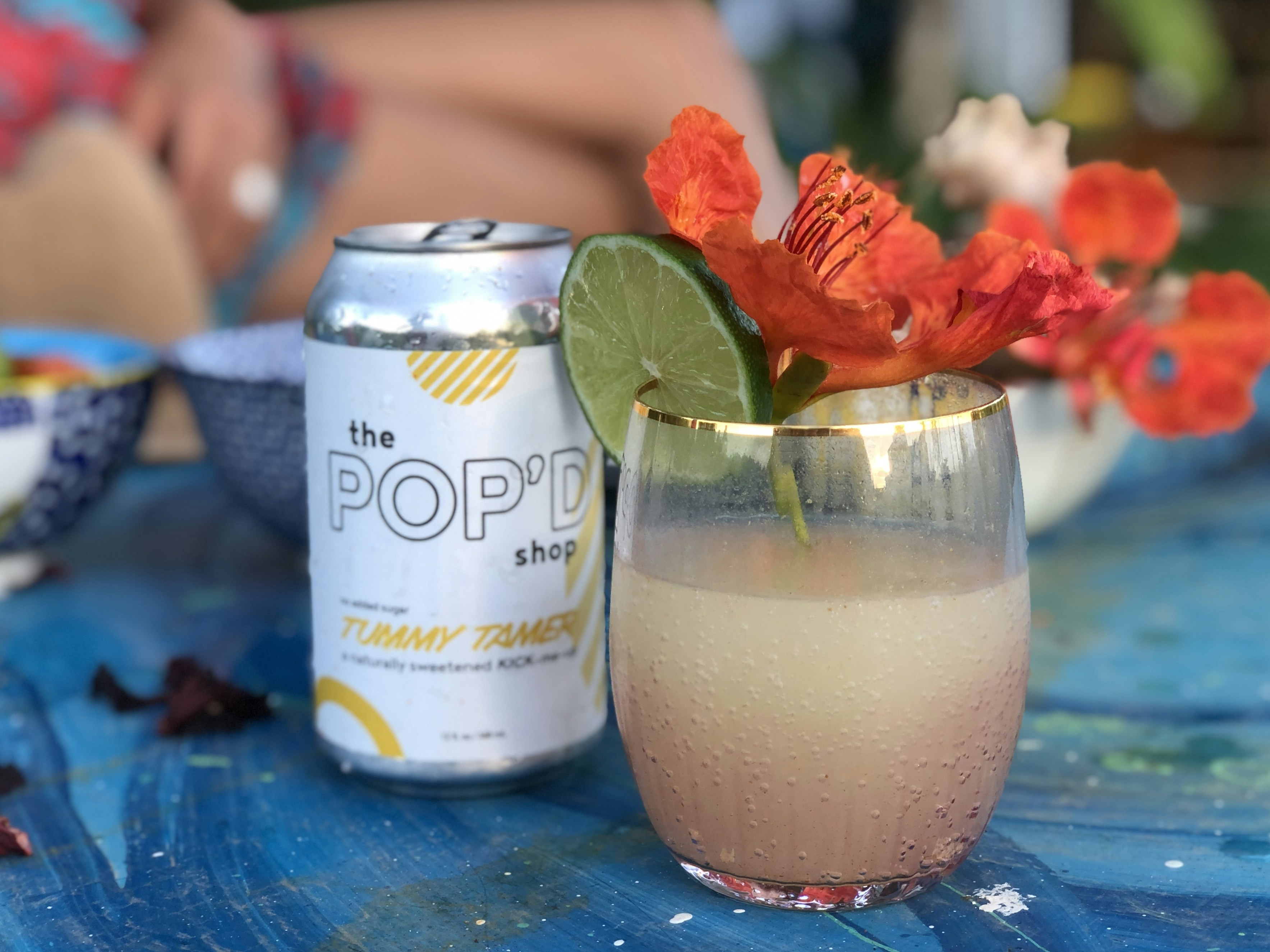 A vibrant cocktail garnished with a bright flower and lime slice, placed on a colorful table. A can of flavored soda sits nearby, enhancing the summery vibe.