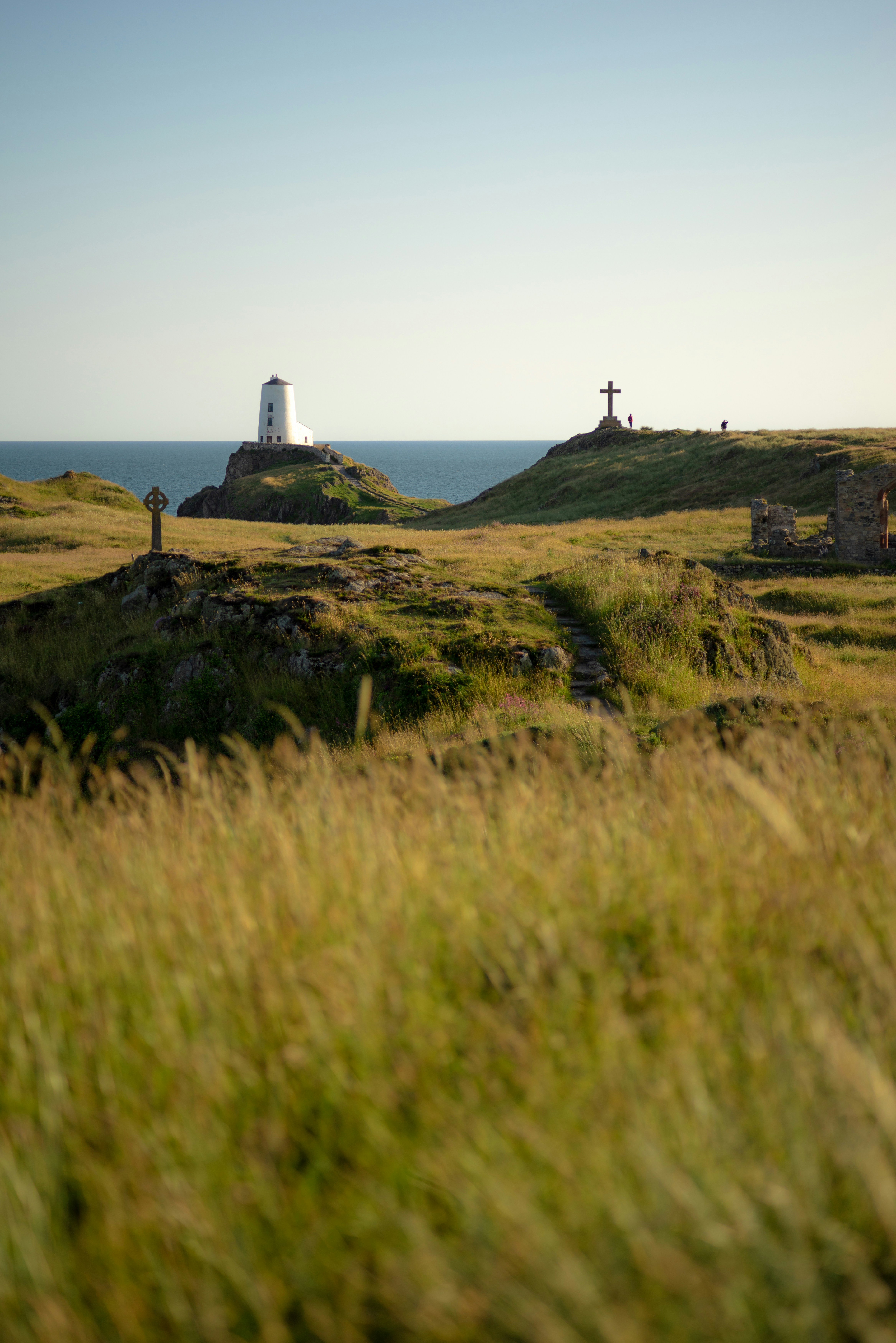 A serene coastal landscape featuring a lighthouse on a rocky outcrop, with ancient crosses dotting the grassy hills under a clear sky.