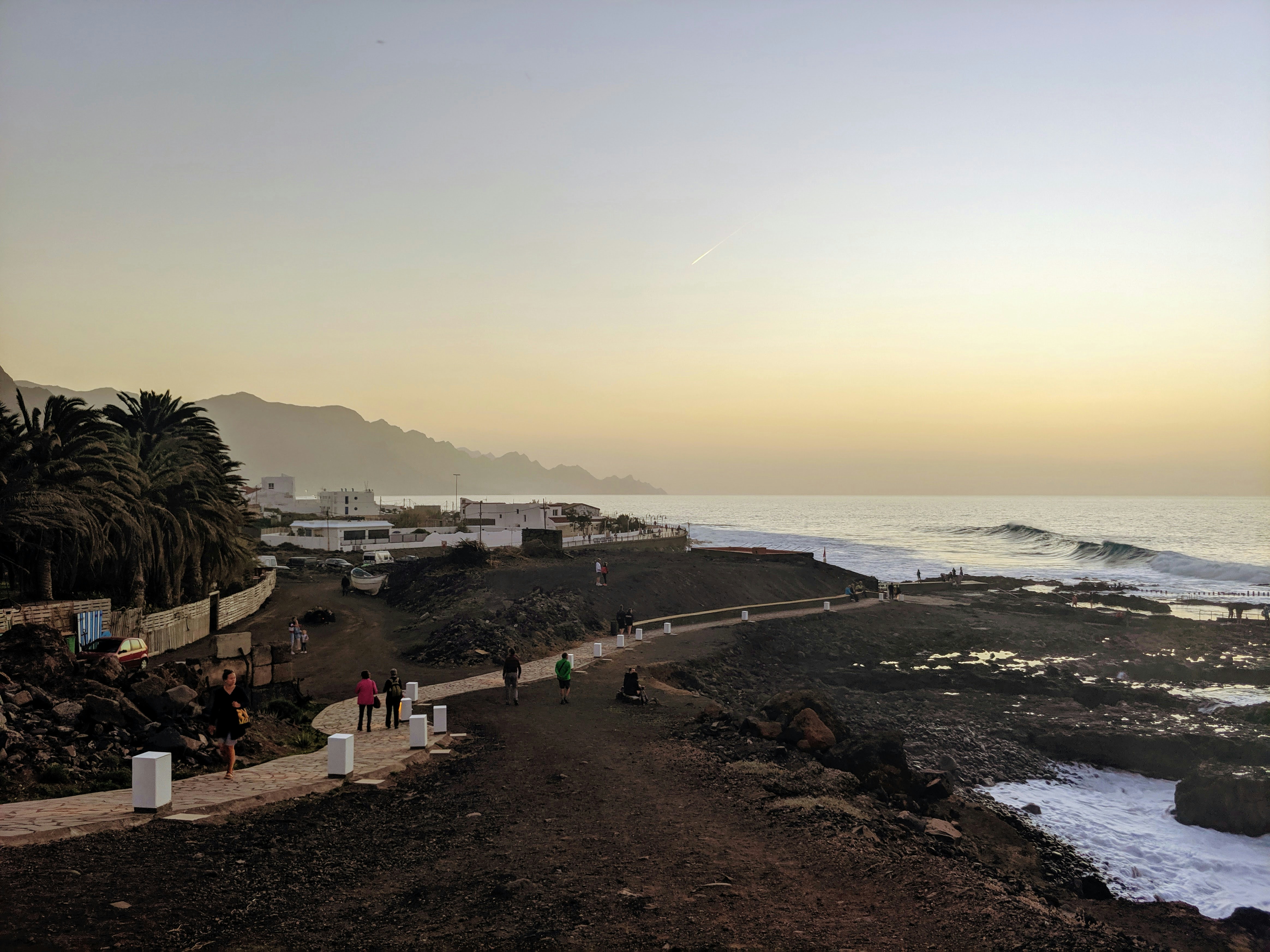 Sunset colors wash over a rocky, lava-dotted coast as a dirt path threads along the shore. Palm silhouettes line the left while the calm sea and distant hills stretch to the horizon.
