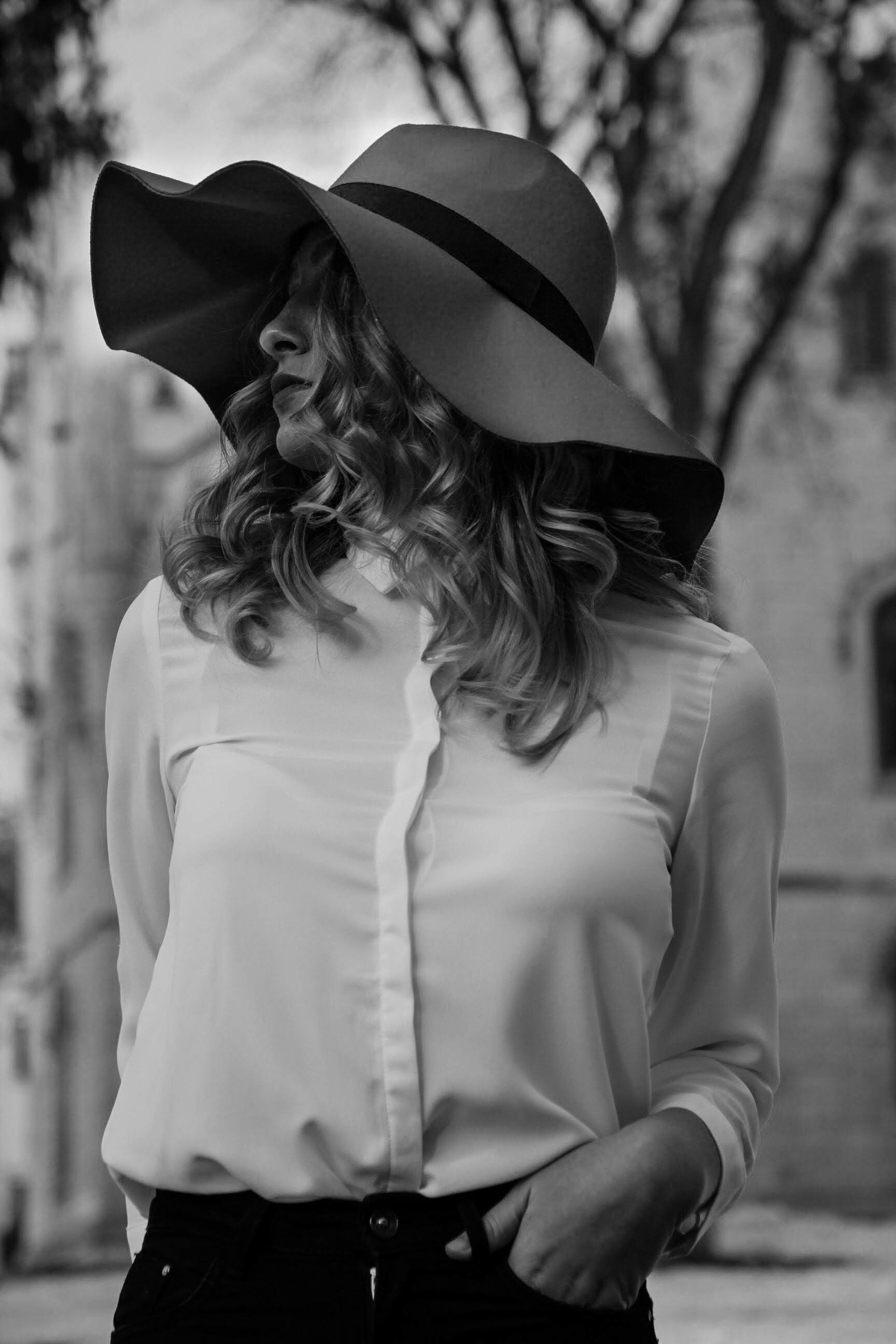 A woman in a wide-brimmed hat gazes away, her curly hair cascading over a light blouse against a blurred architectural backdrop.