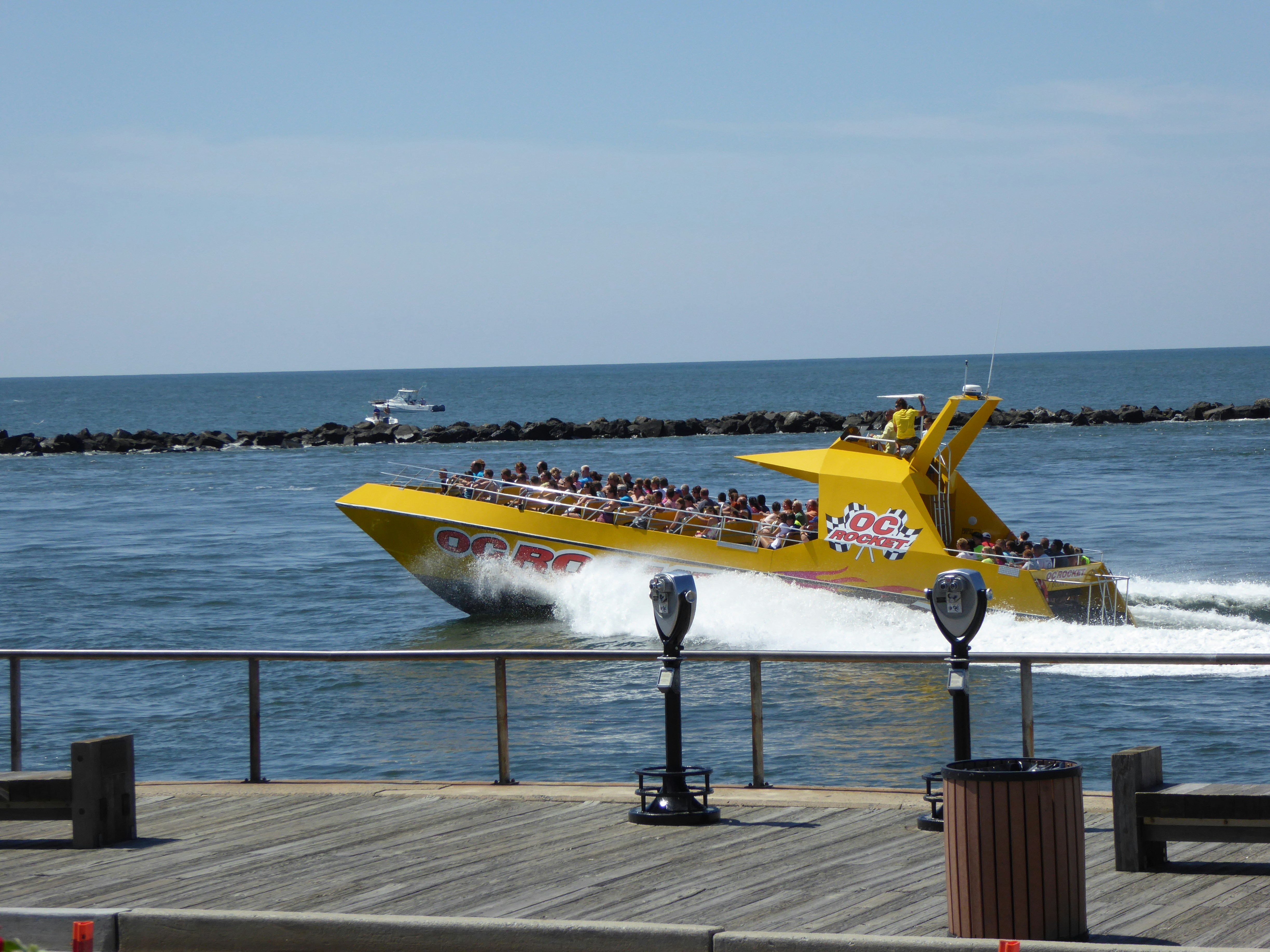 A vibrant yellow jet boat speeds across the water, carrying excited passengers, with a calm sea and distant boat in the background.