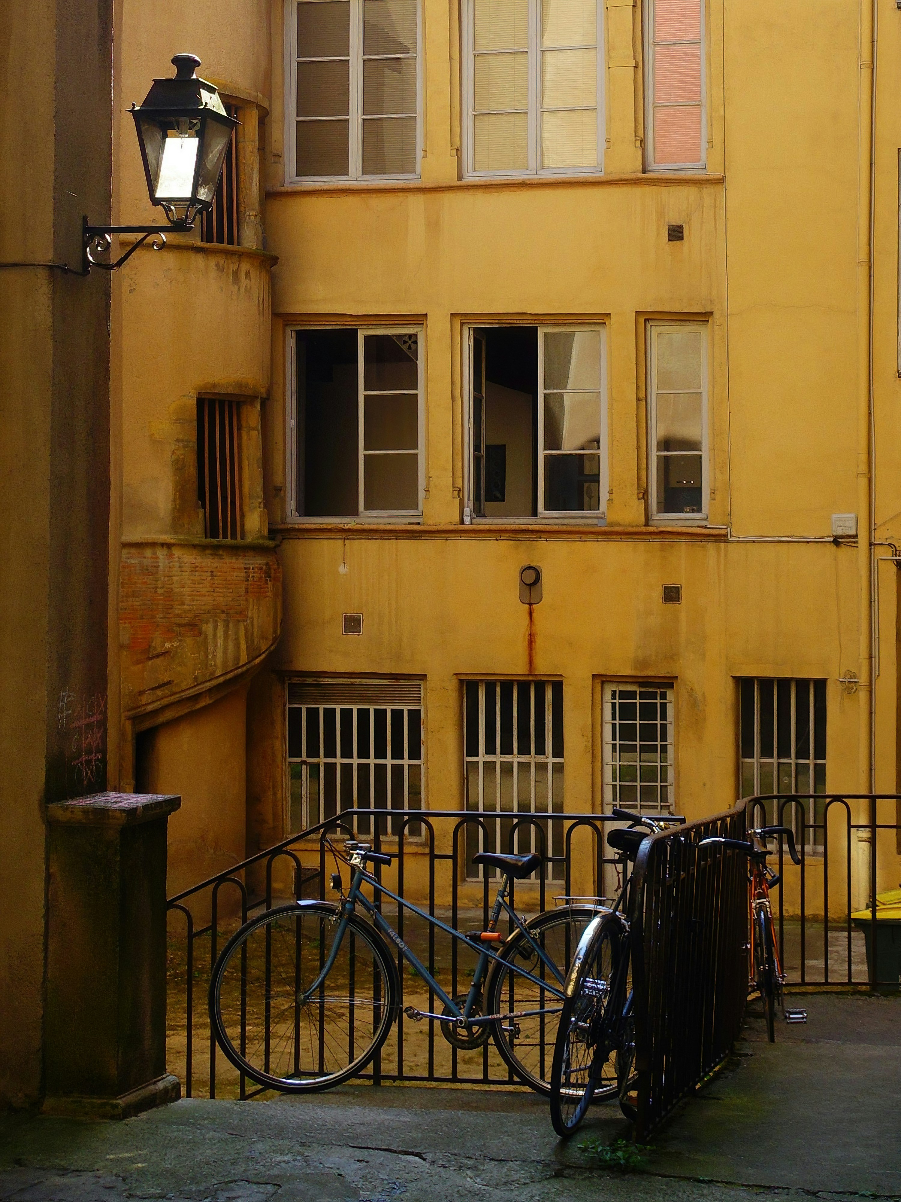 A tranquil courtyard scene featuring bicycles parked against a wrought-iron railing, surrounded by warm-toned buildings with large windows.