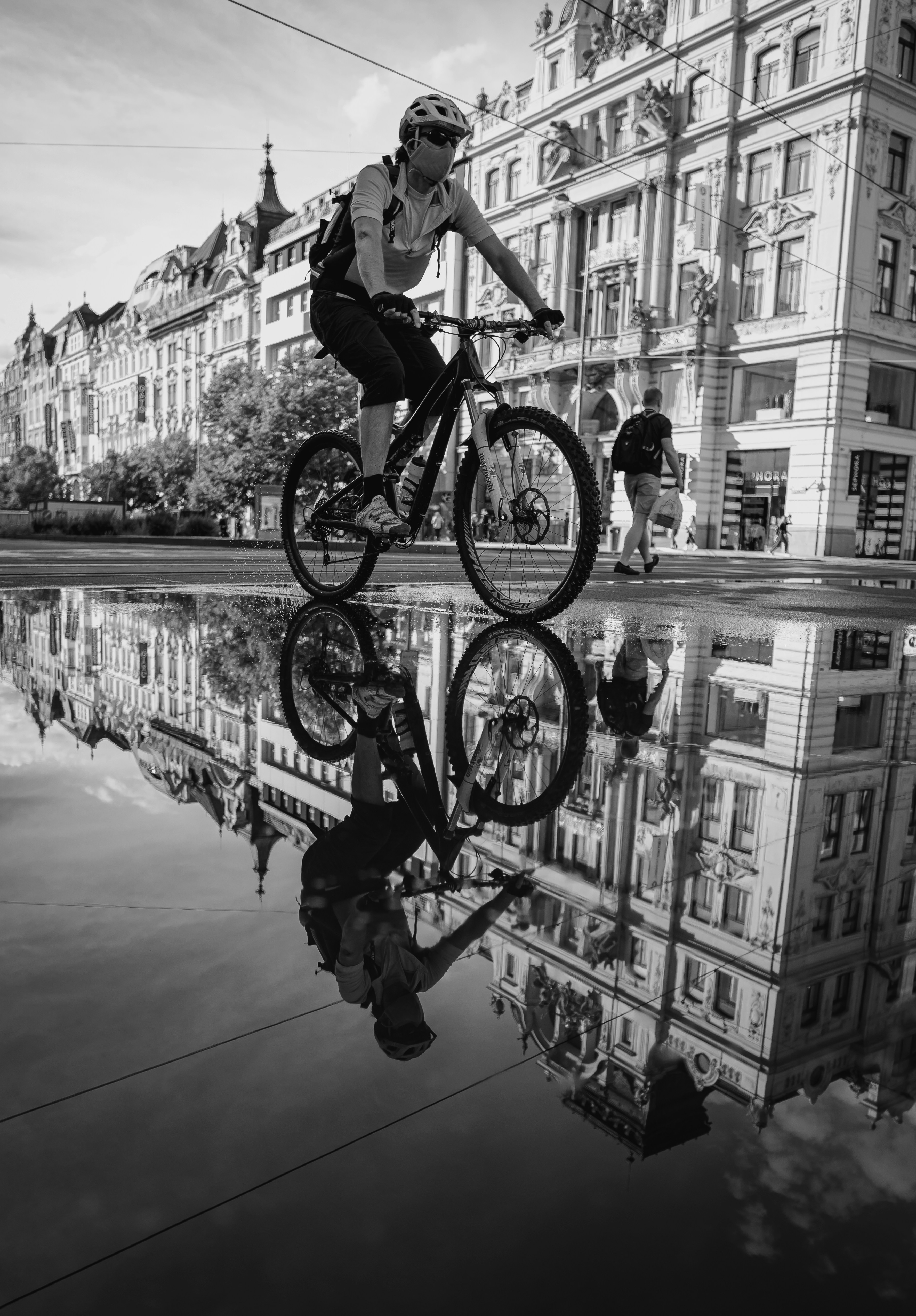 Cyclist navigating a city street, reflected in a puddle that mirrors the historic architecture around. Black and white tones enhance the urban atmosphere.