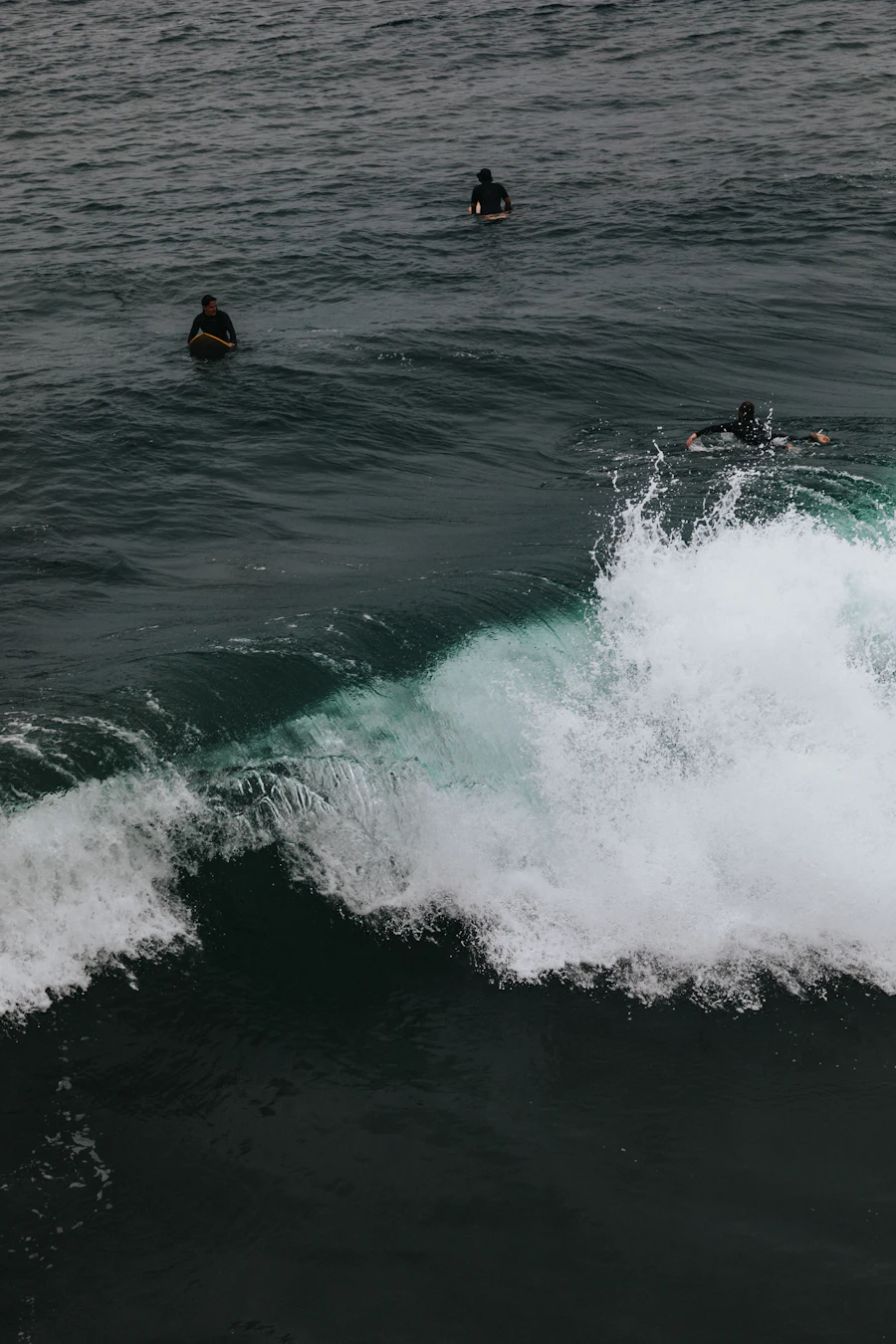 Surfers waiting in the ocean, poised for the next wave, as a crashing wave creates a frothy display. The scene captures the thrill of surfing in dynamic waters.