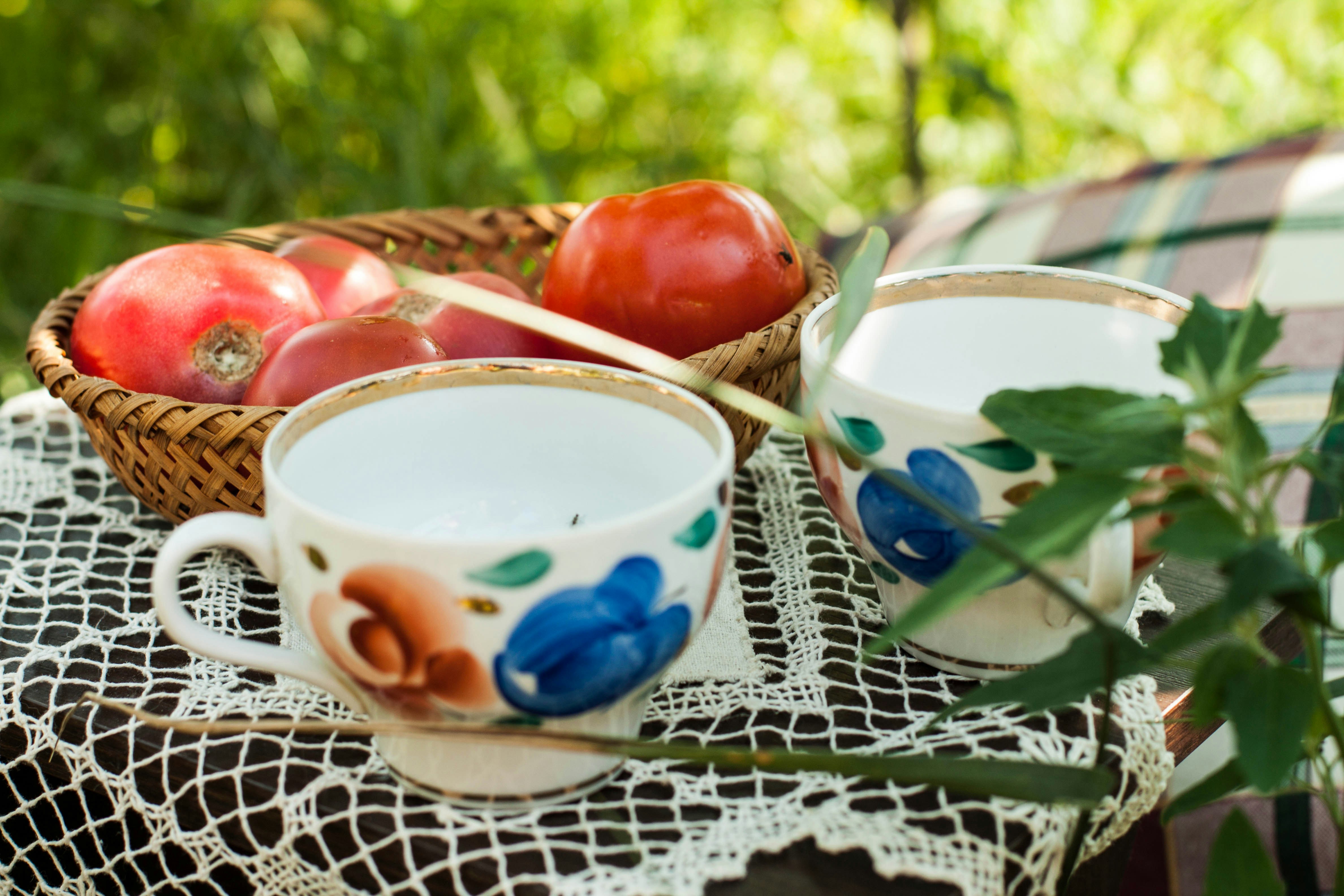 Delicate porcelain cups sit on a lace tablecloth beside a woven basket filled with ripe tomatoes, surrounded by lush greenery.