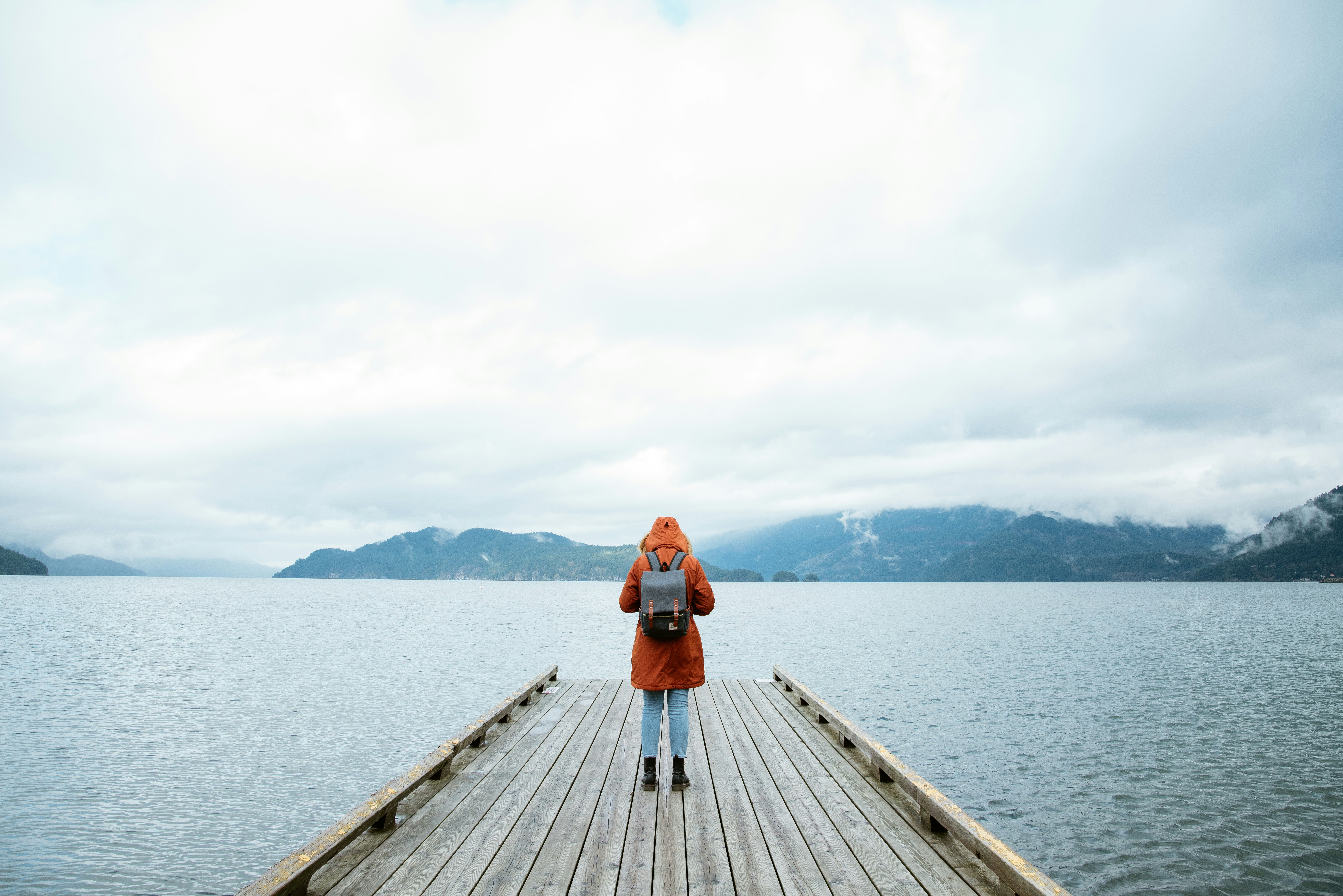 Orange-Coated Wanderer at the Pier's Edge Over a Misty Fjord