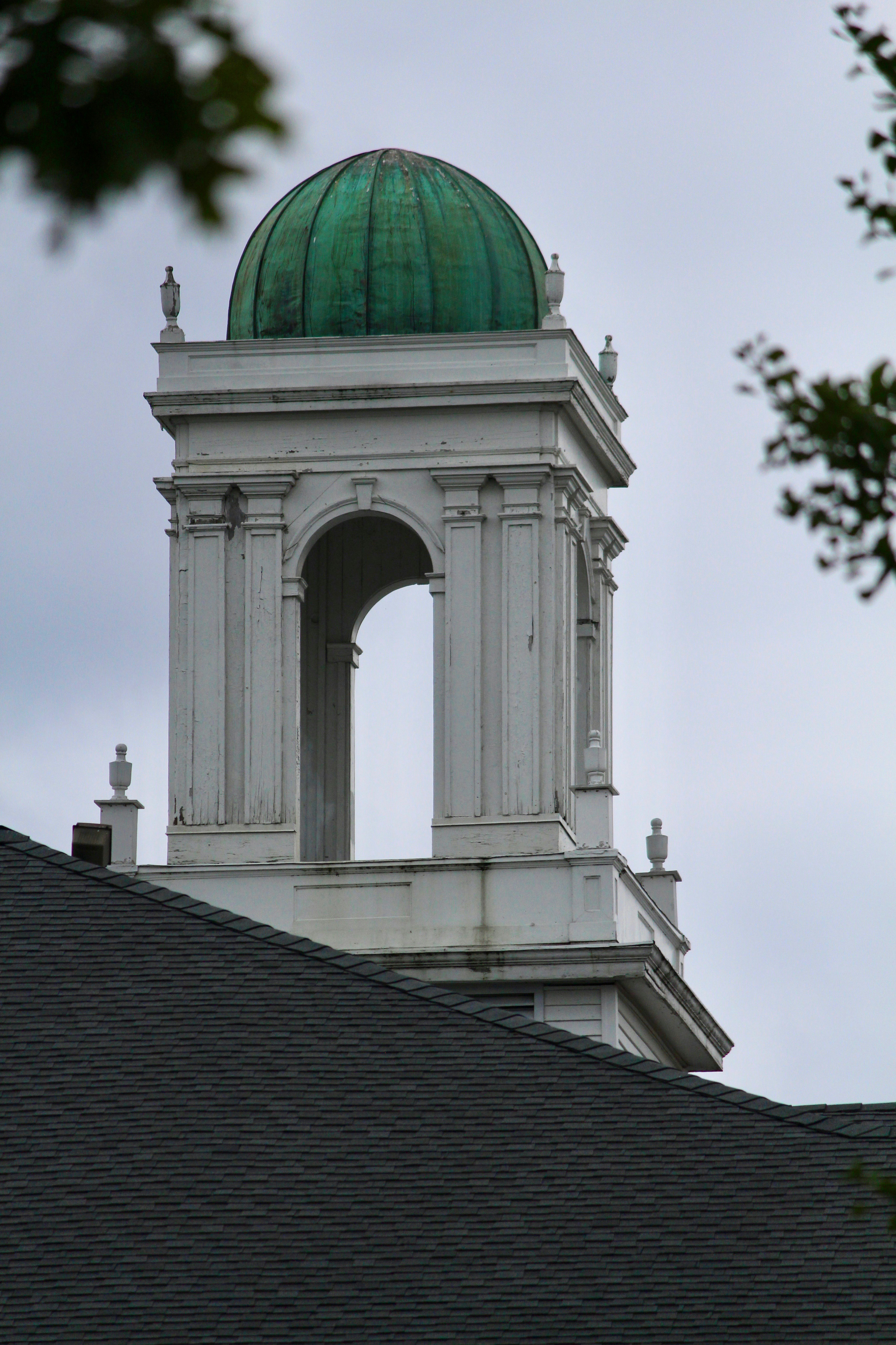 Architectural tower with a green dome peeking above a sloped roof, showcasing intricate details against a cloudy sky.