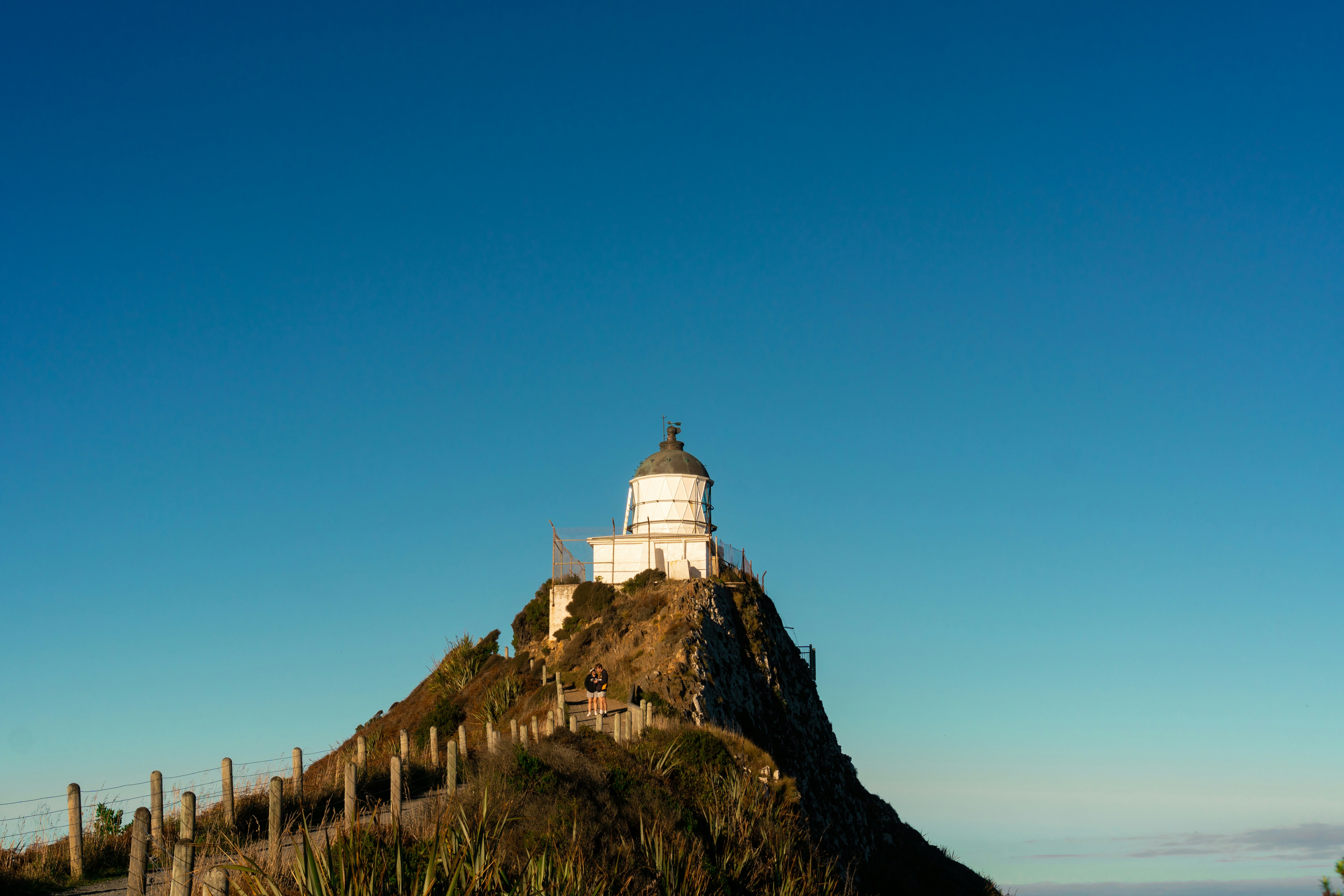 Historic lighthouse perched on a rugged hill, surrounded by lush greenery and a clear blue sky. The structure stands as a testament to maritime navigation.